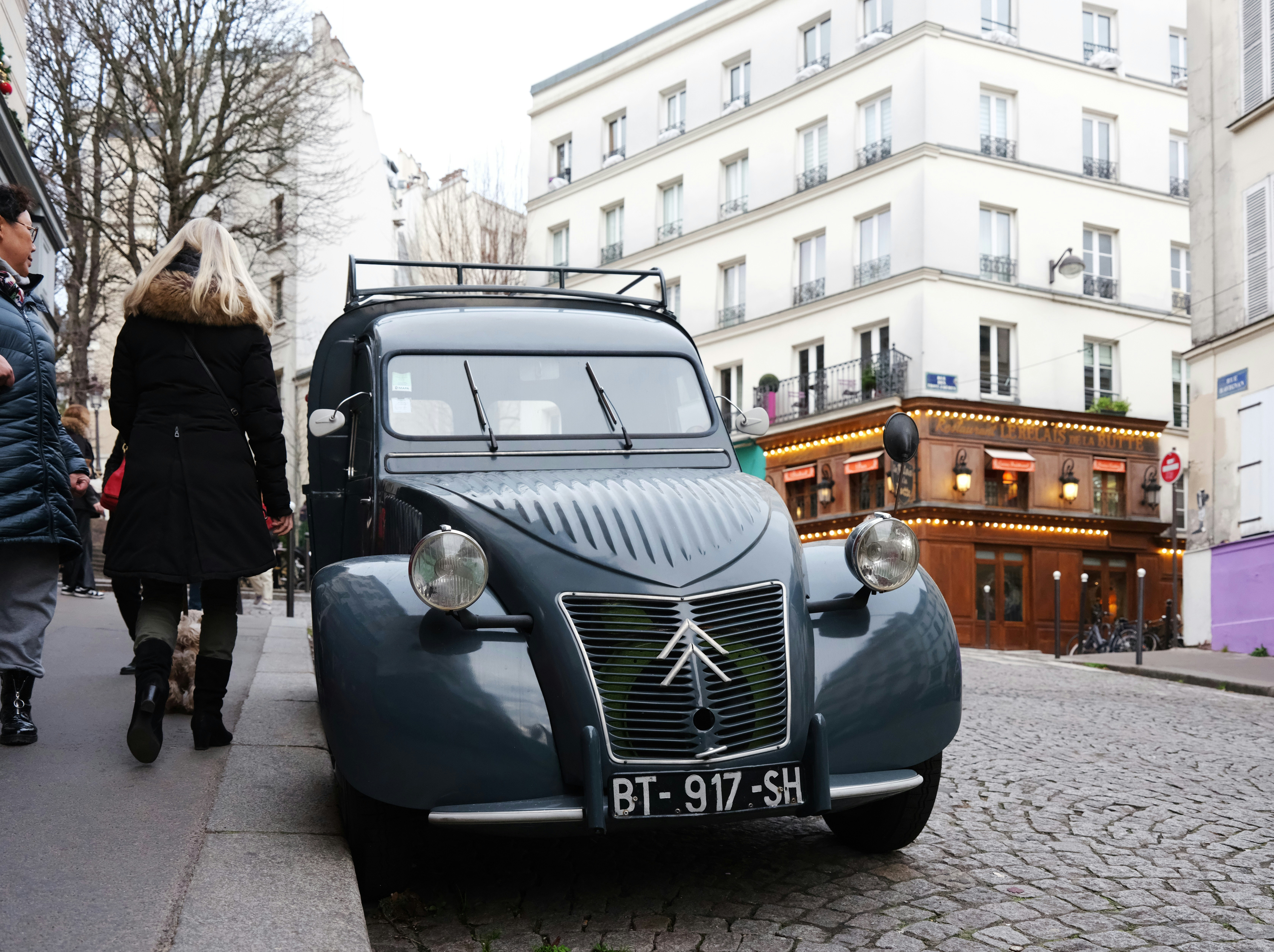 A vintage French car sits on a cobblestone street with pedestrians nearby and a warmly lit café in the background.