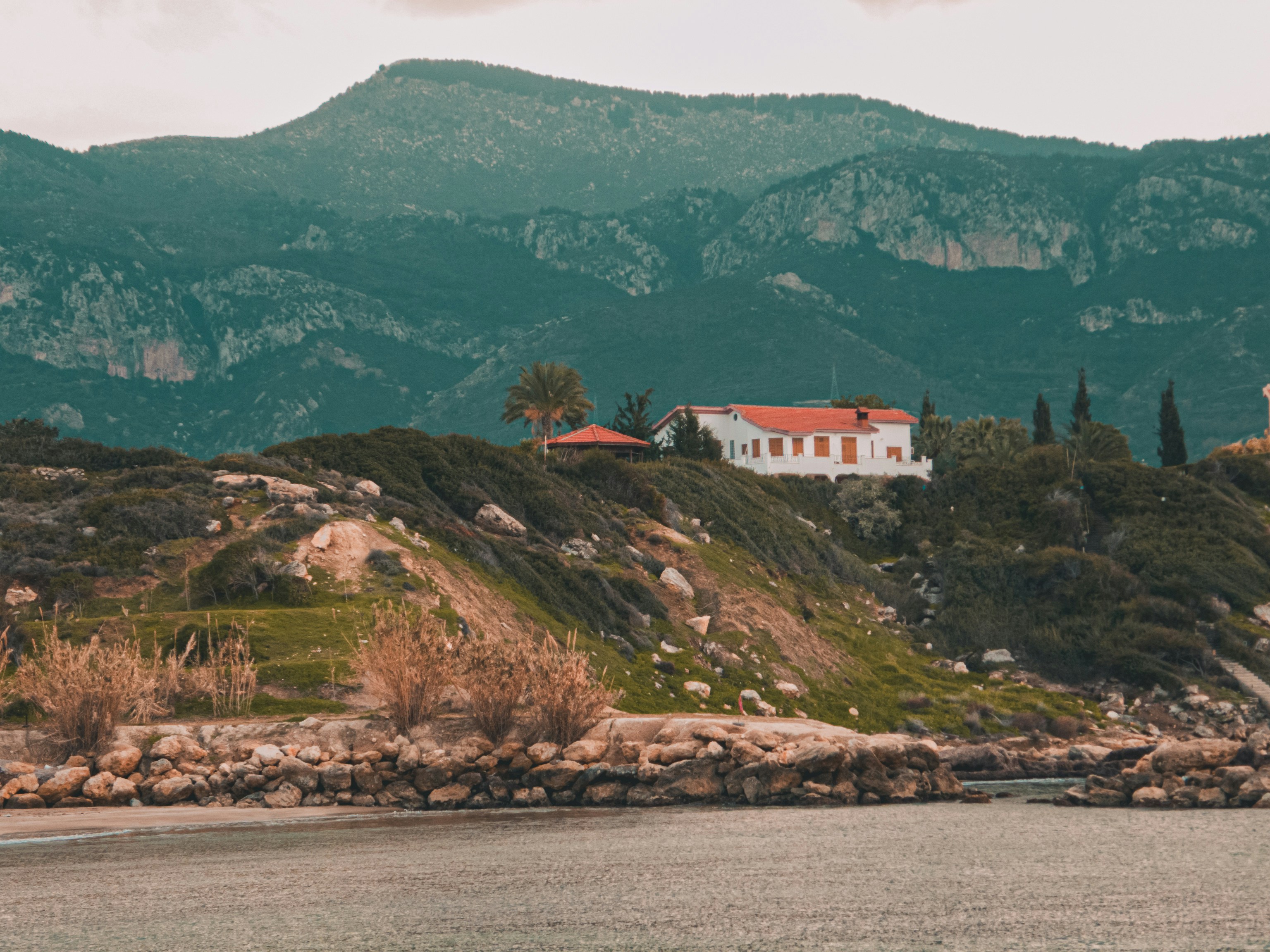 Coastal hillside photograph featuring a white building with a red roof perched above a rocky shoreline, with distant green mountains providing a dramatic backdrop.