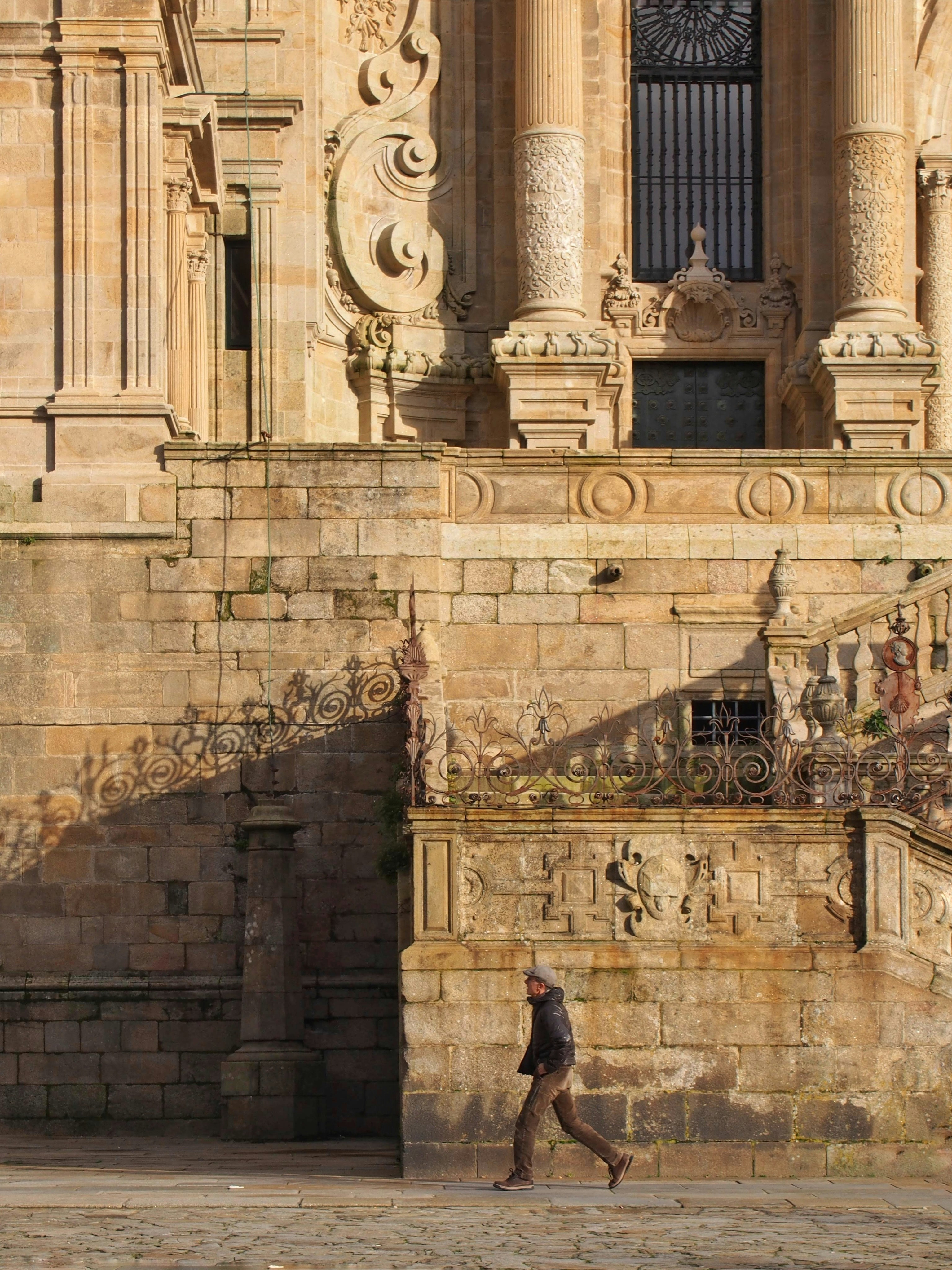 Photograph capturing the sun-washed façade and ornate railing as a lone figure walks along the warm, stone terrace.
