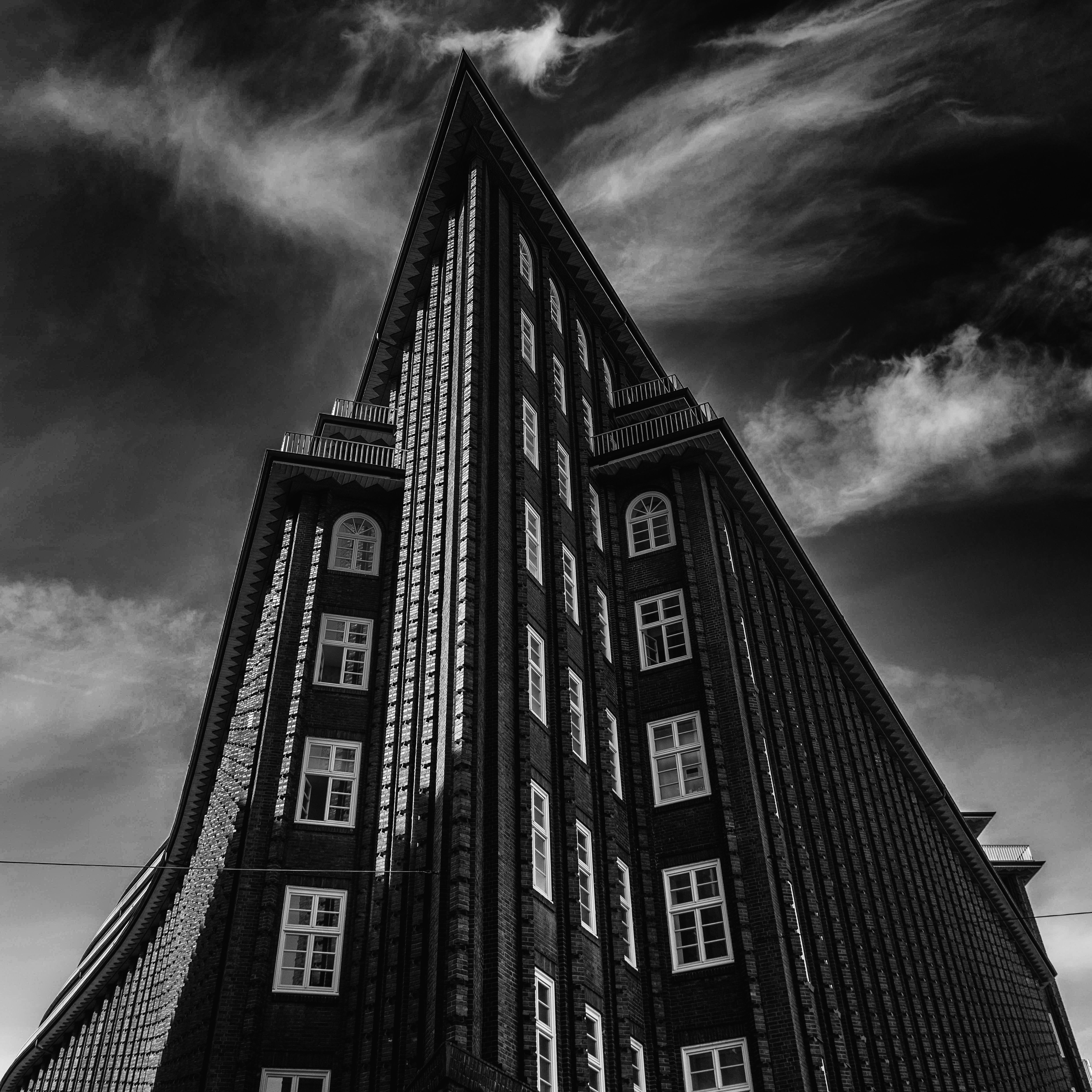 Monochrome exterior of a tall, sharply angled brick building rising toward dramatic clouds, emphasizing perspective and geometry. A structural landmark image suitable for architectural study.