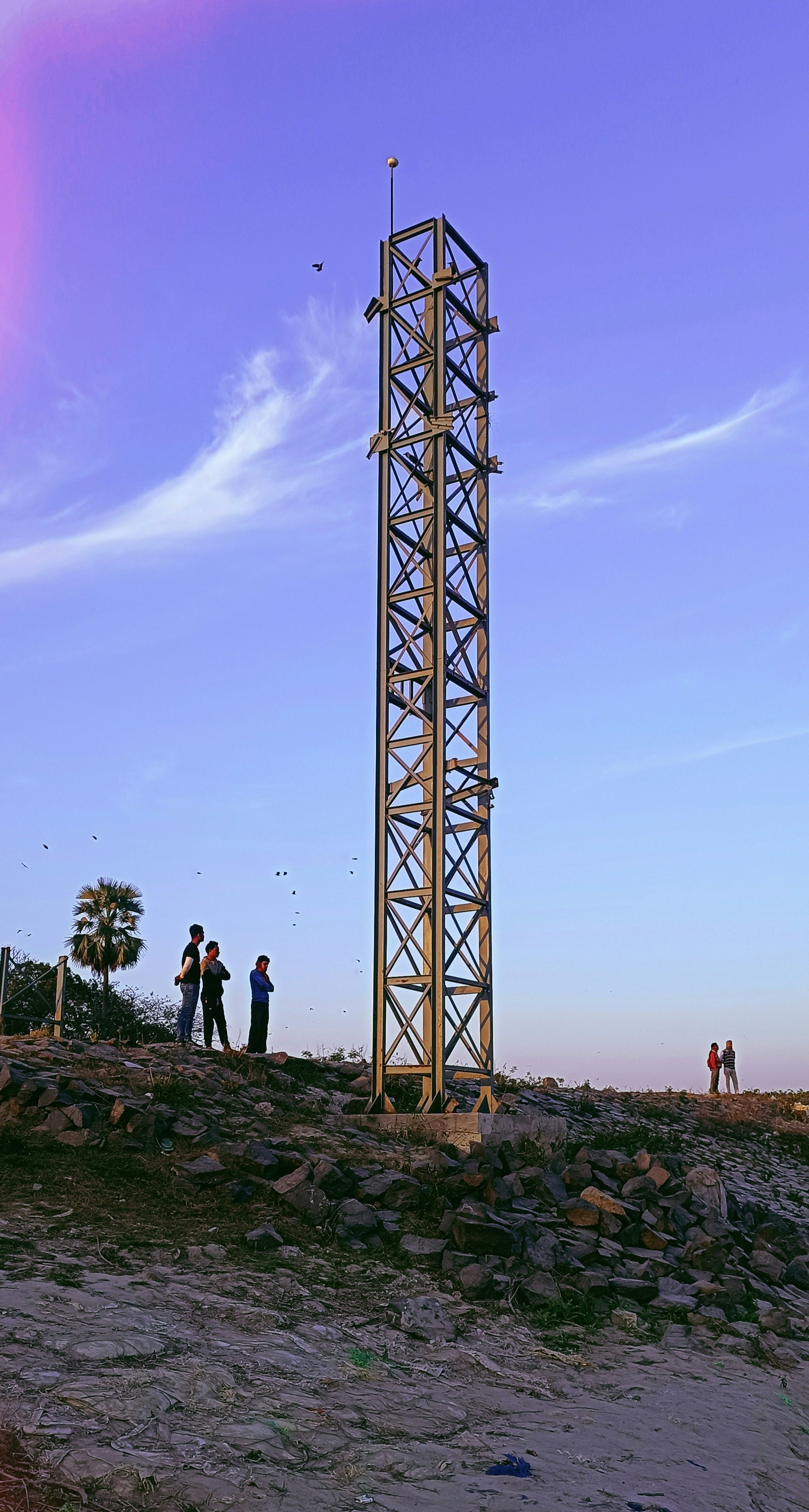 Crane-like lattice tower rises against a pastel sky with a group of people on rocky shore at dusk; a lone couple stands farther away, adding depth to the scene. (Photo/survey of a coastal area, with a clear sky.)