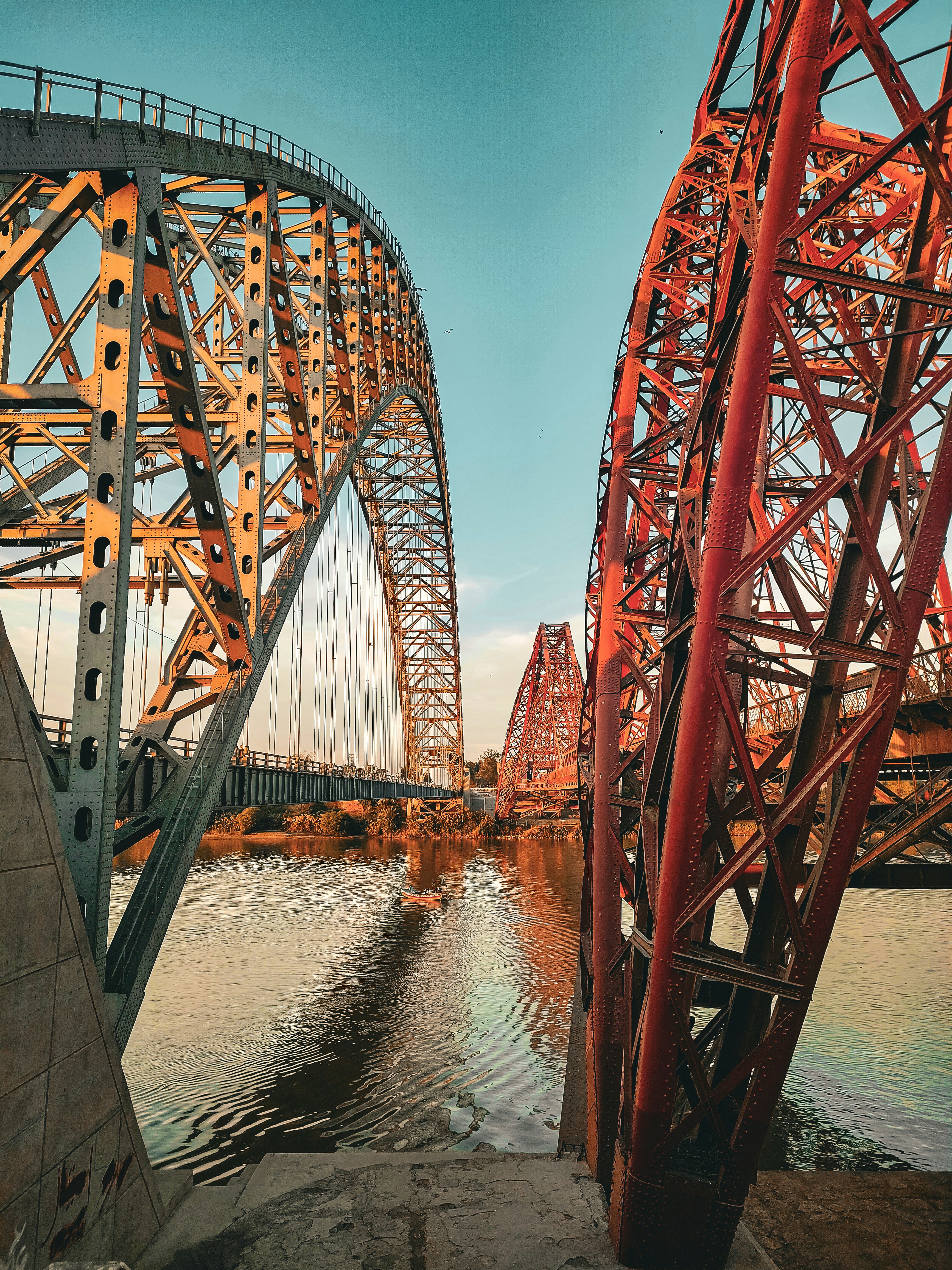 A photograph of rusted red and steel bridge arches spanning a calm river at dusk. The composition emphasizes perspective lines and reflections along the water.