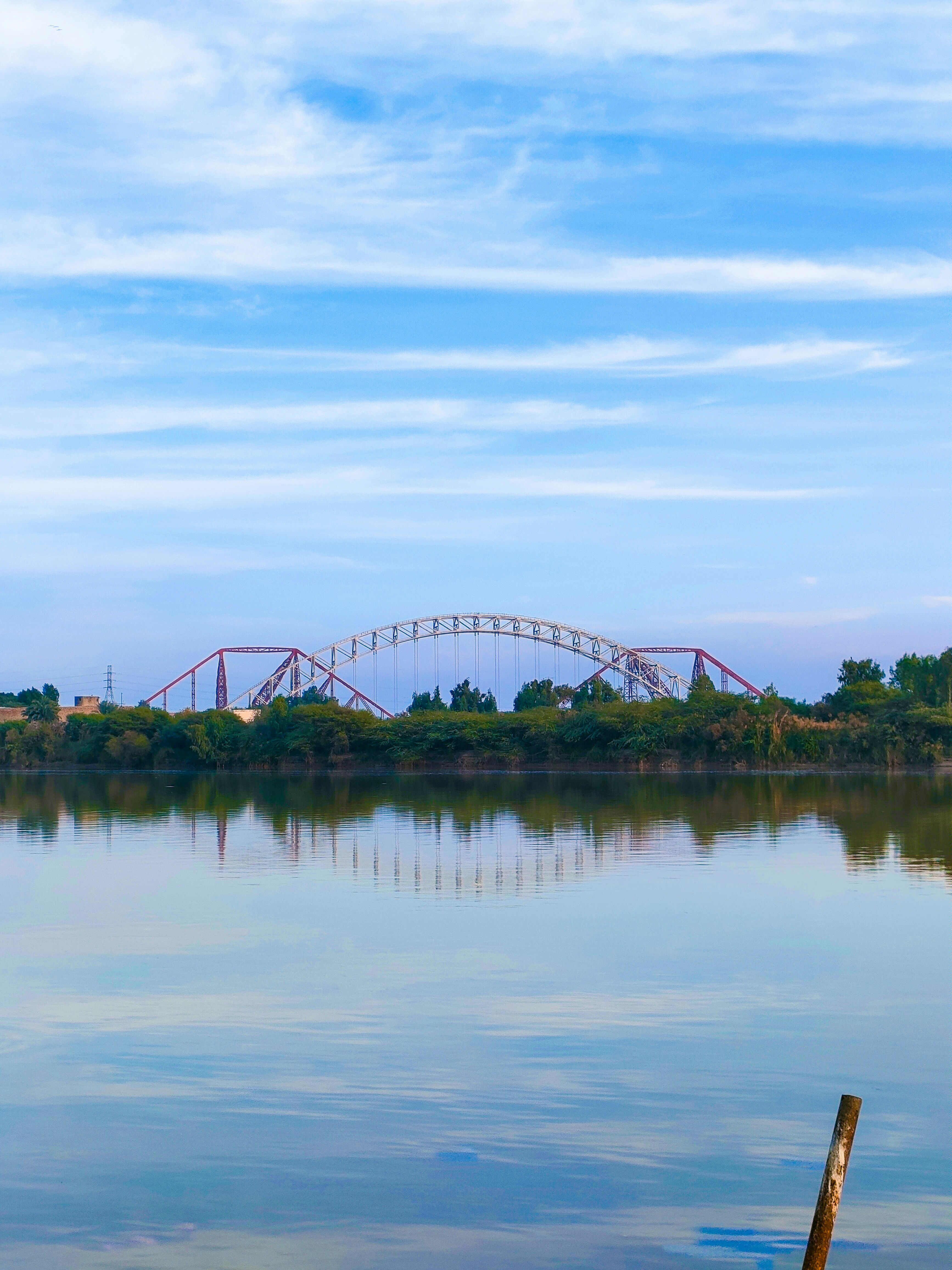 A tranquil riverside scene featuring a distant arched bridge rising above the tree line, with a calm water surface reflecting the sky and structure. Photograph highlights a serene landscape with soft hues and sky gradients.