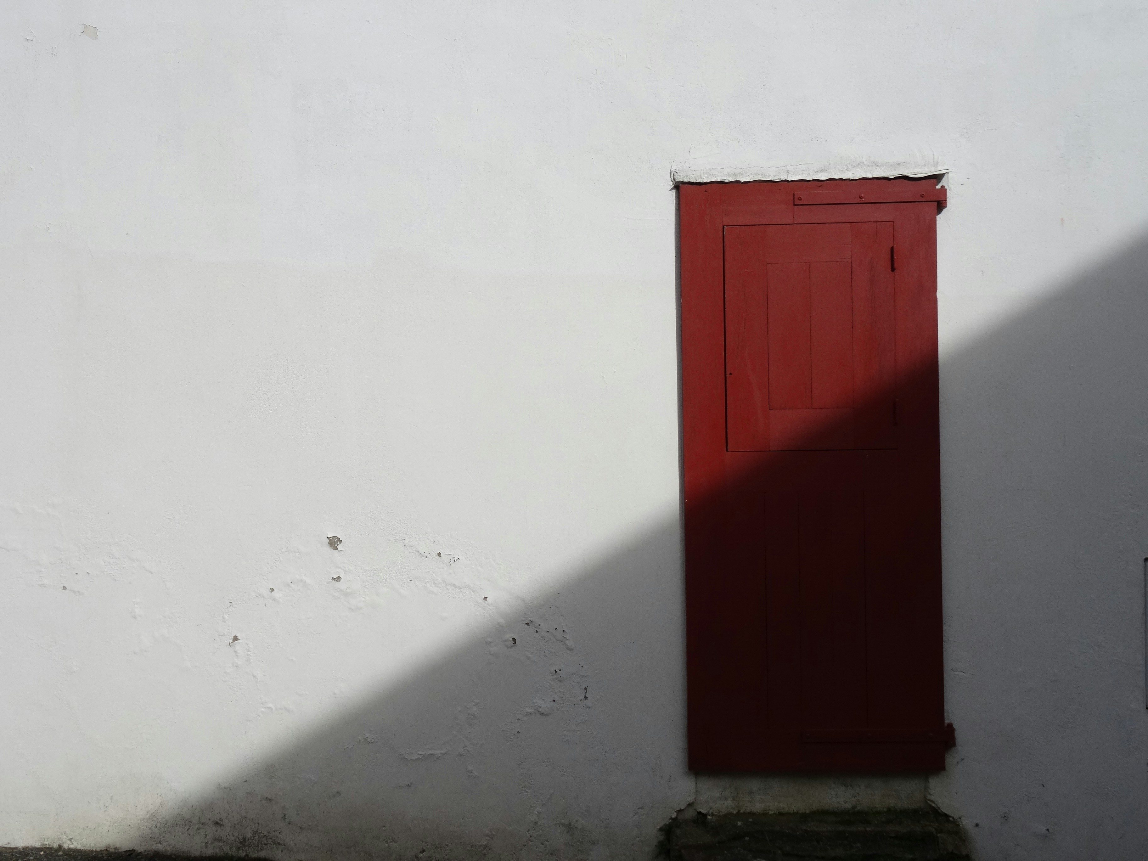 A white stucco wall with a right-side red door, bathed in a sharp diagonal shadow that slices across the surface.