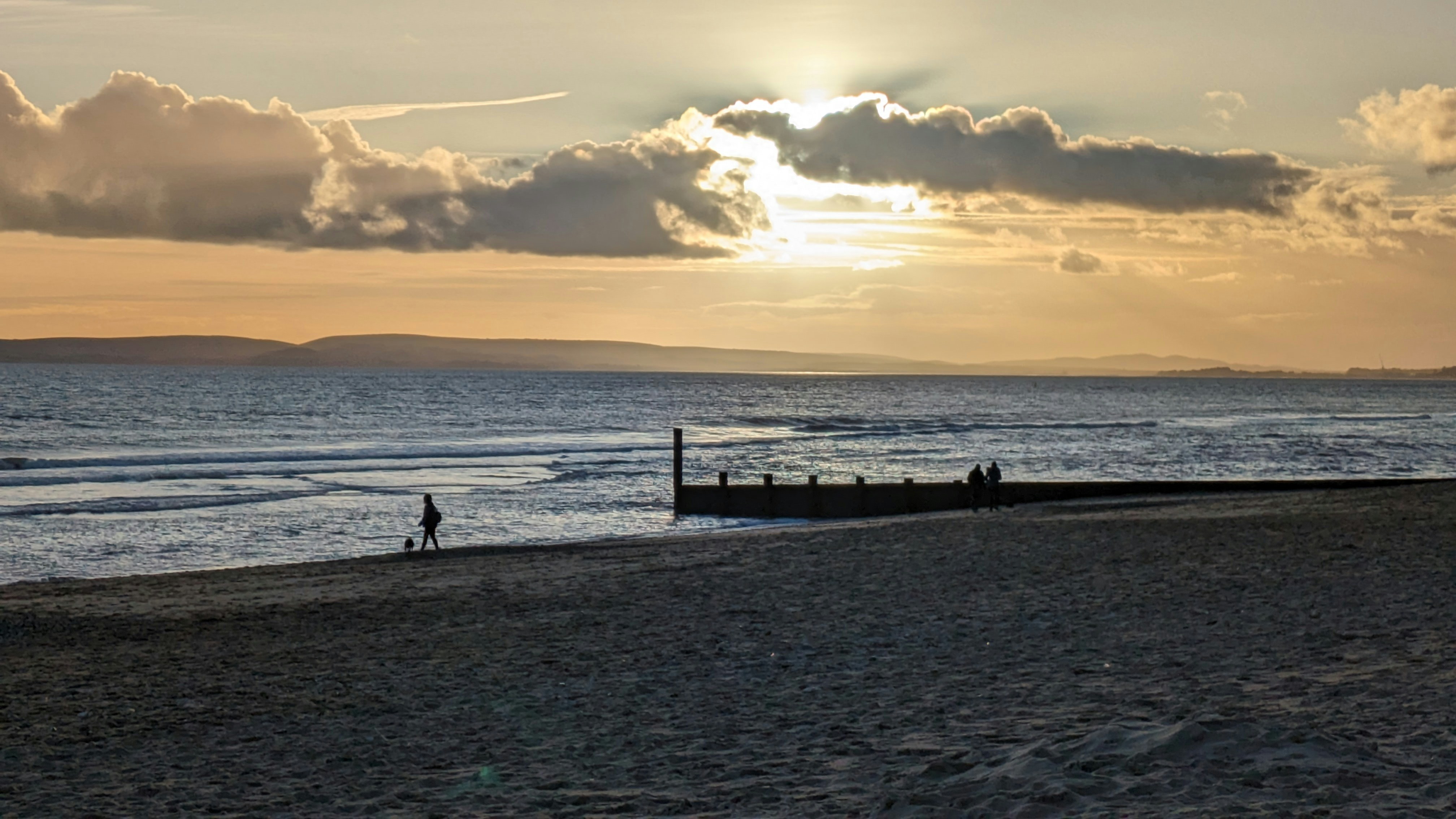 Silhouetted figures on a sandy beach under a dramatic sunset sky with distant hills.