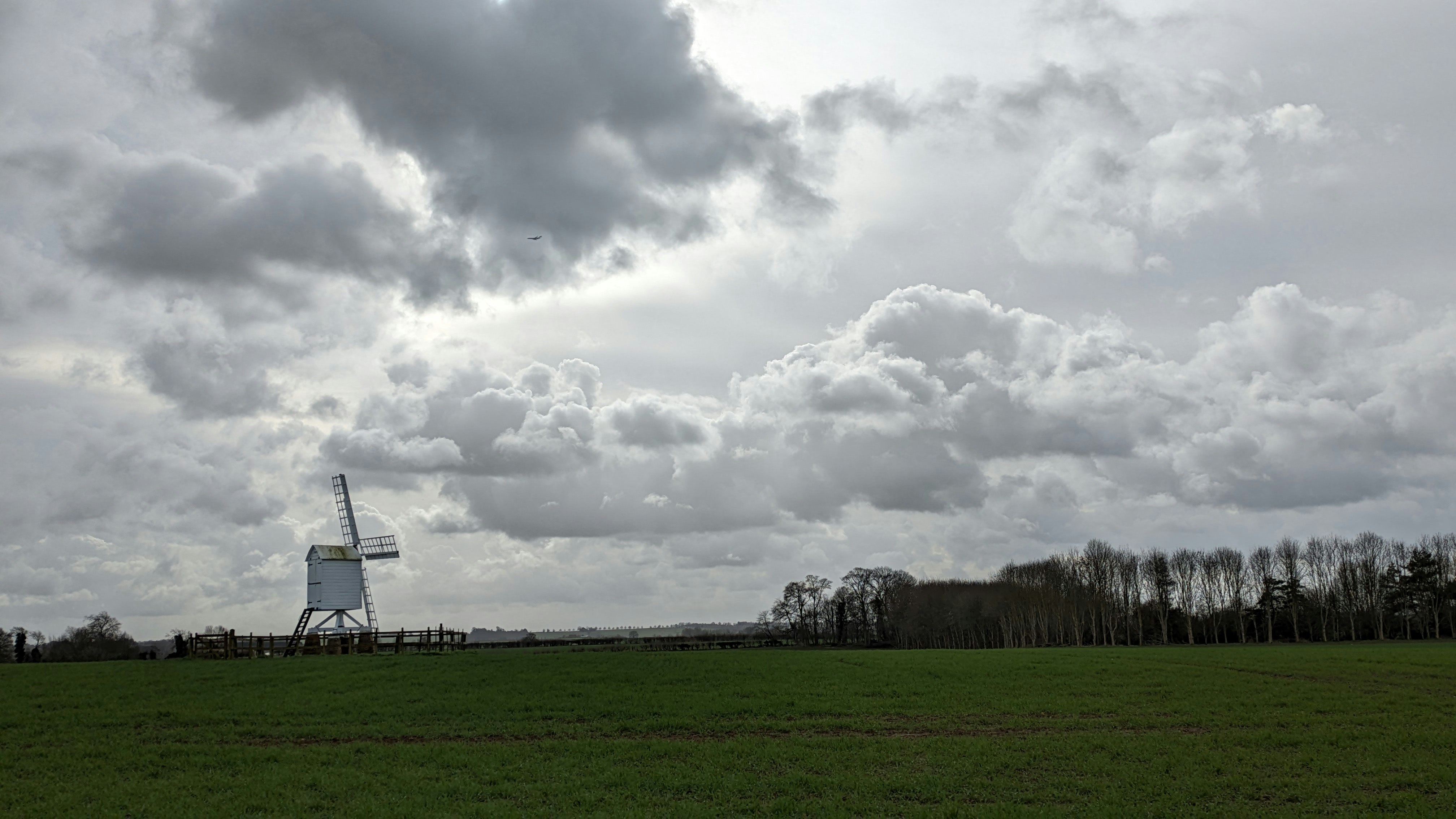 Windmill on a grassy field beneath a cloud-filled sky, with a distant line of trees on the horizon. The composition emphasizes expansive rural space.
