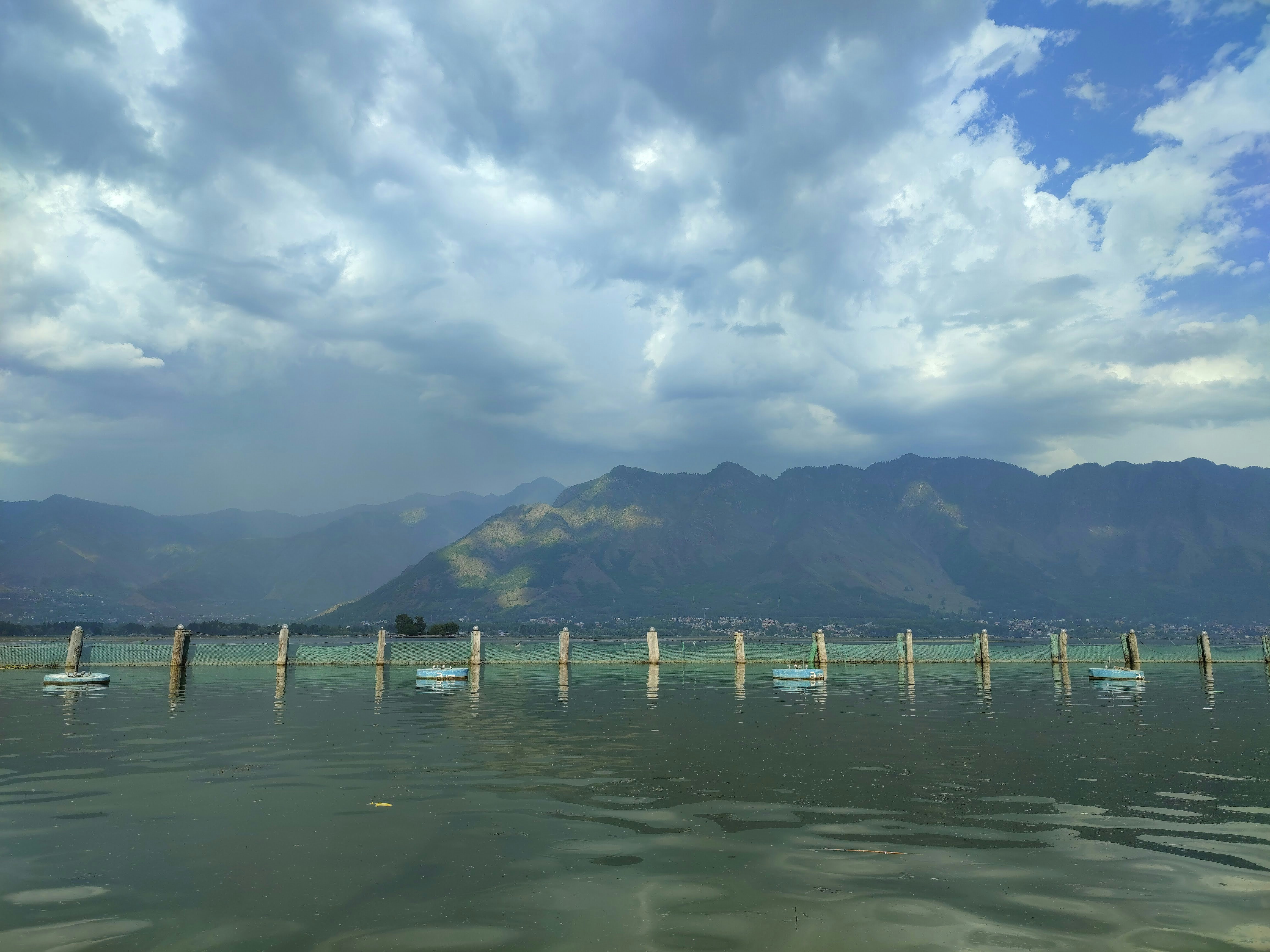Wooden posts emerge from a calm lake, with mountains and dramatic clouds in the background.