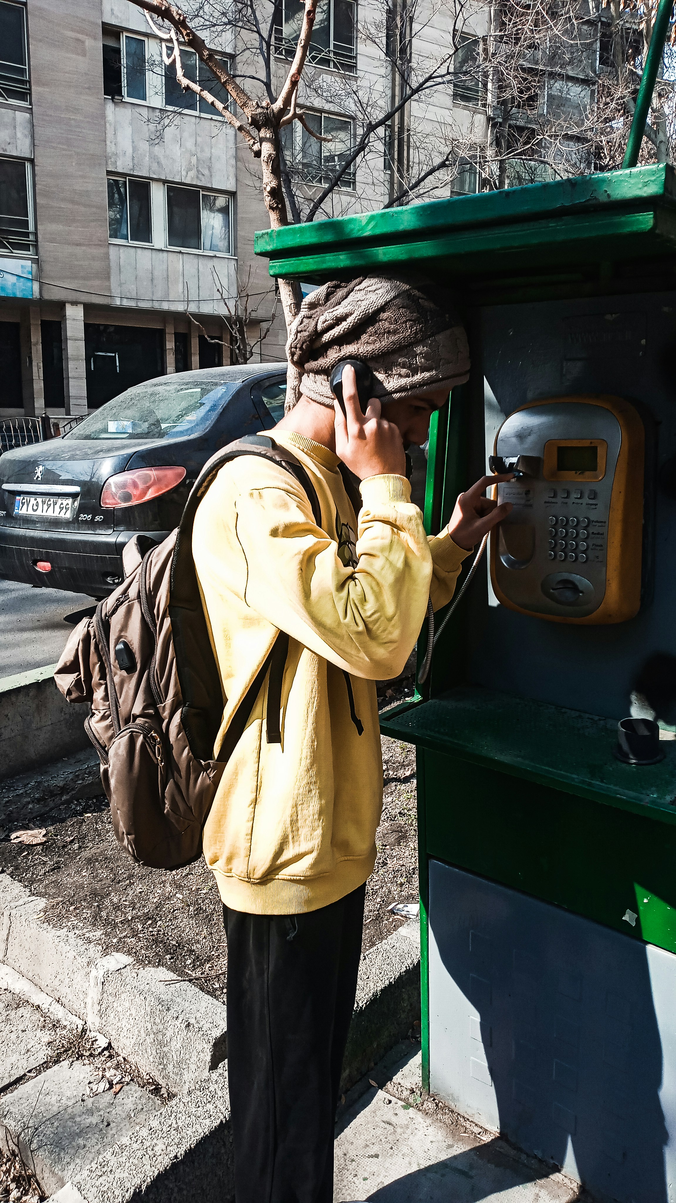 Young man in a yellow hoodie, backpack on his back, speaks on a public phone at a green booth on a city street; a parked car and apartment building frame the scene.