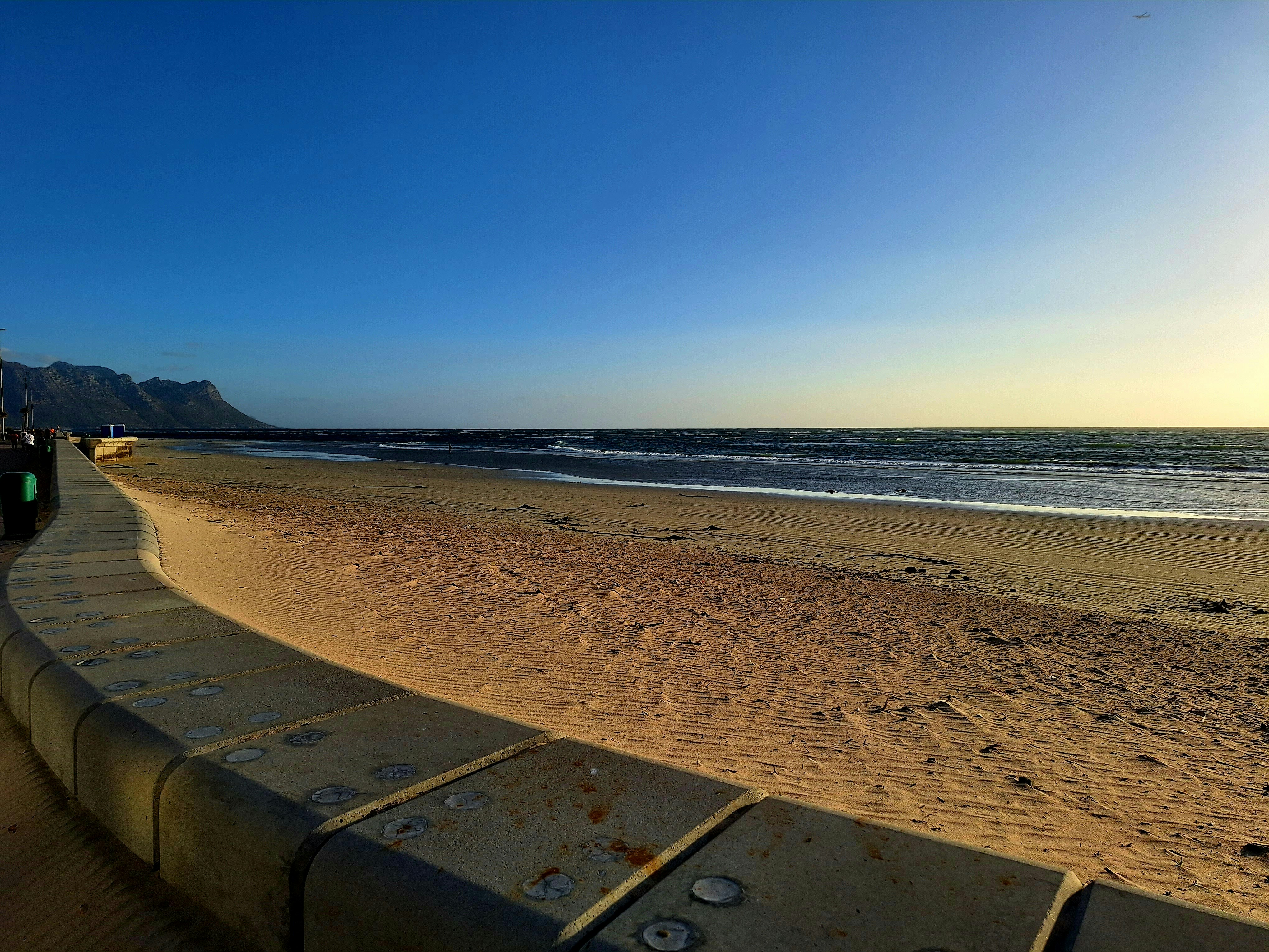 Curved beachfront promenade with gentle waves and mountains under a clear blue sky.