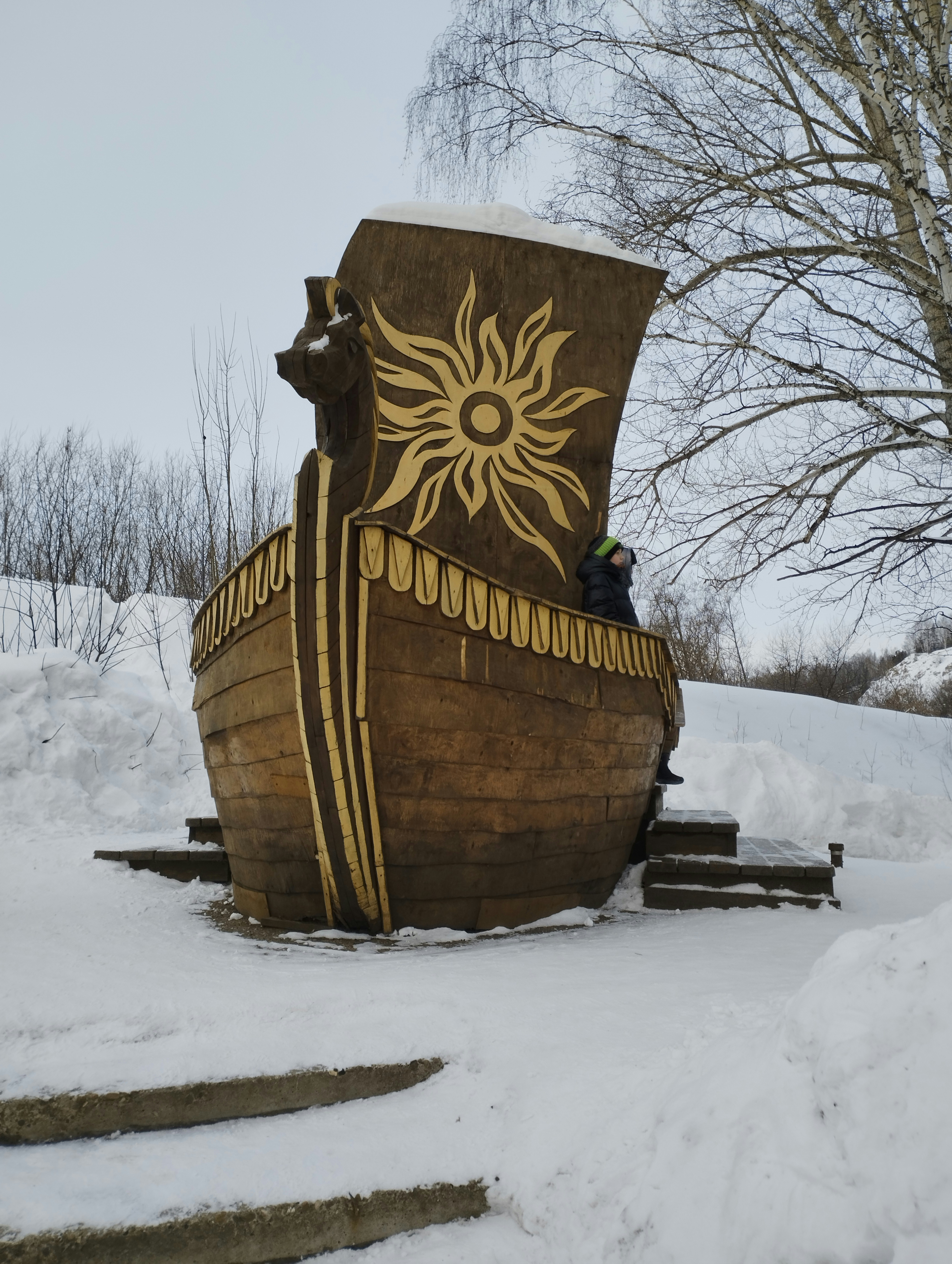 Snow-blanketed wooden Viking ship sculpture with sun motif stands as the focal feature in a wintry park; people in jackets inspect the monument. The scene emphasizes the carved craft against bare trees and snow banks.