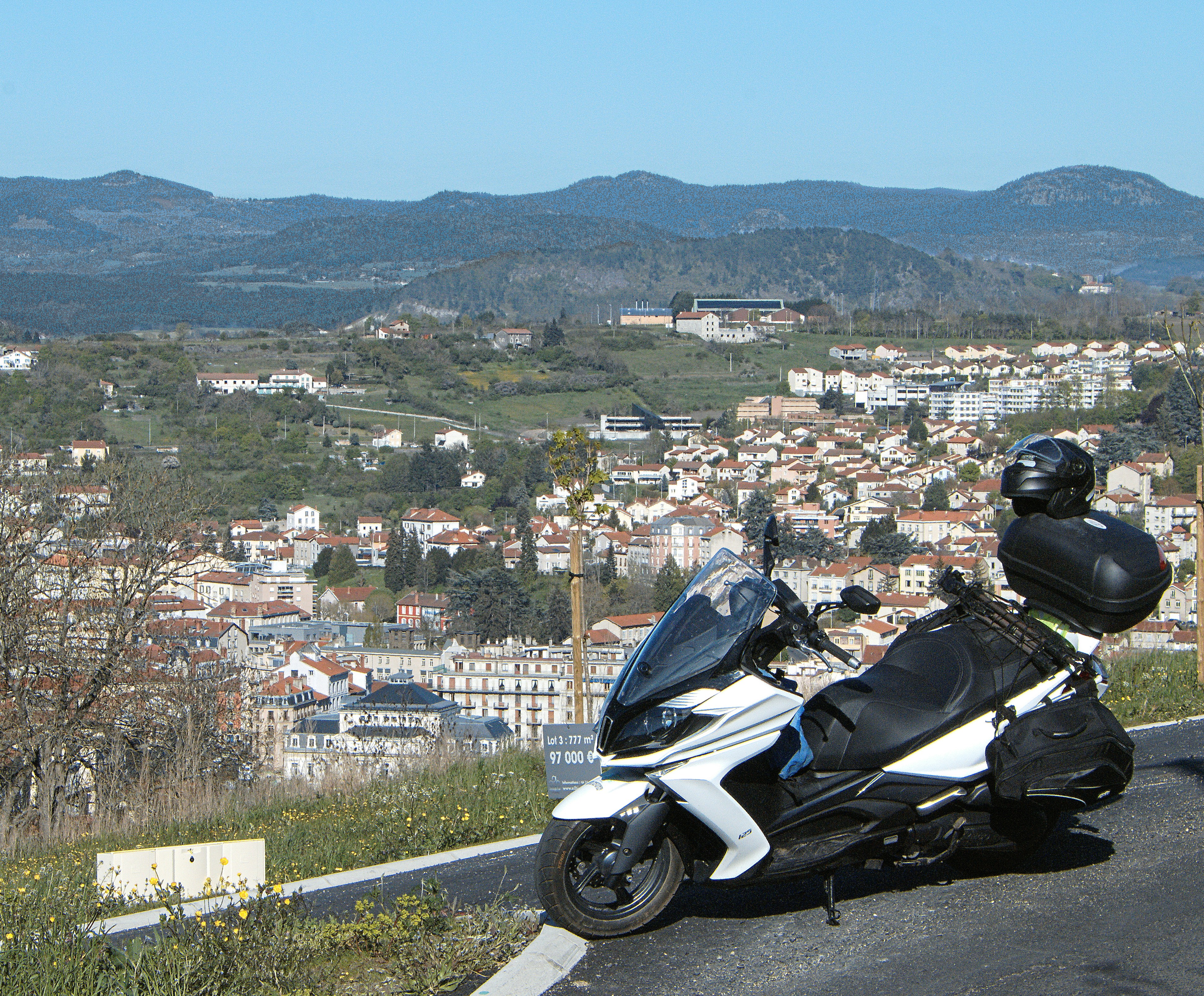 Motorcycle parked on a roadside with a scenic view of a hillside town and mountains under a clear blue sky.