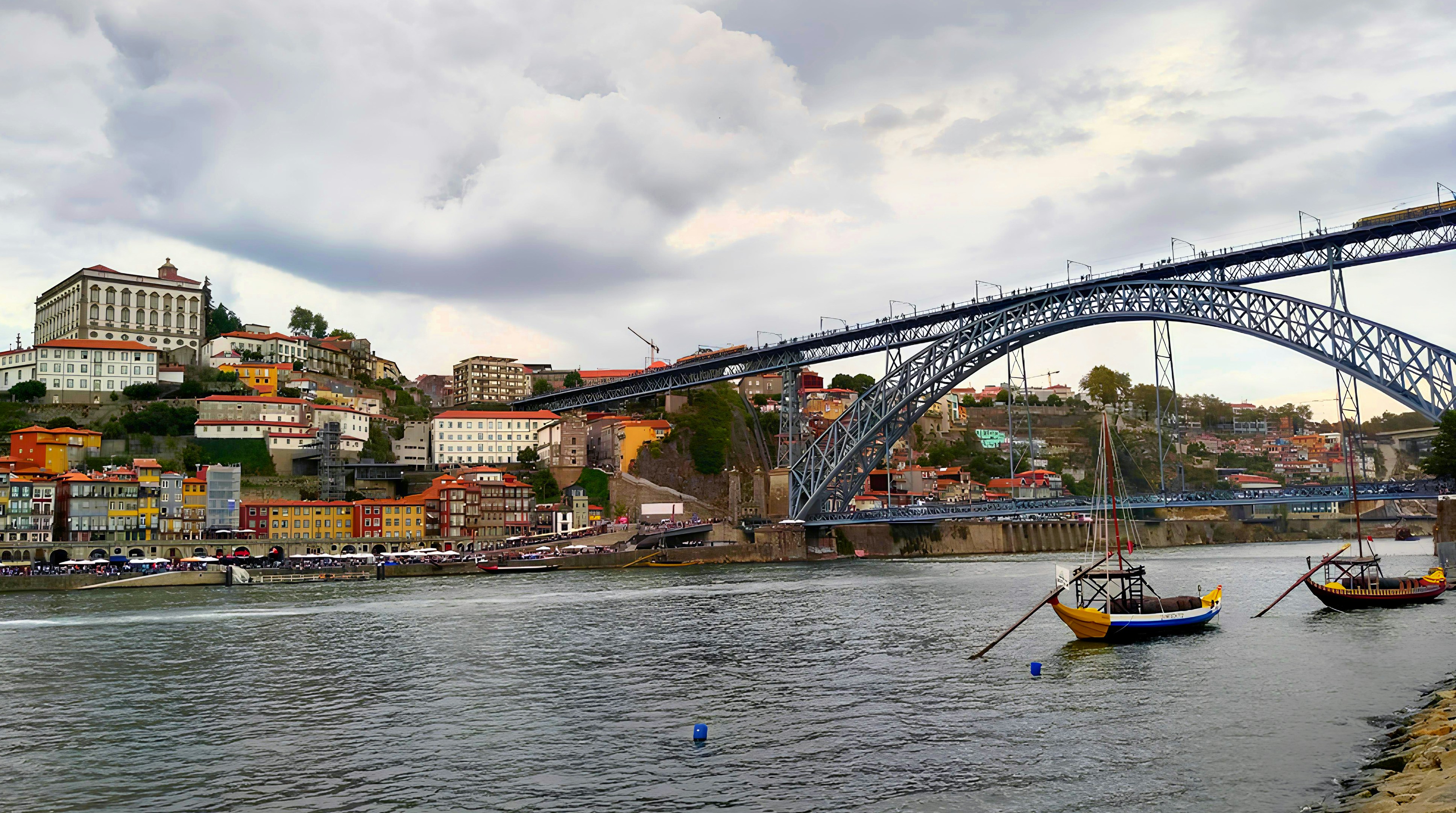 Iconic arched bridge spans the Douro River with colorful hillside buildings in Porto under a cloudy sky.