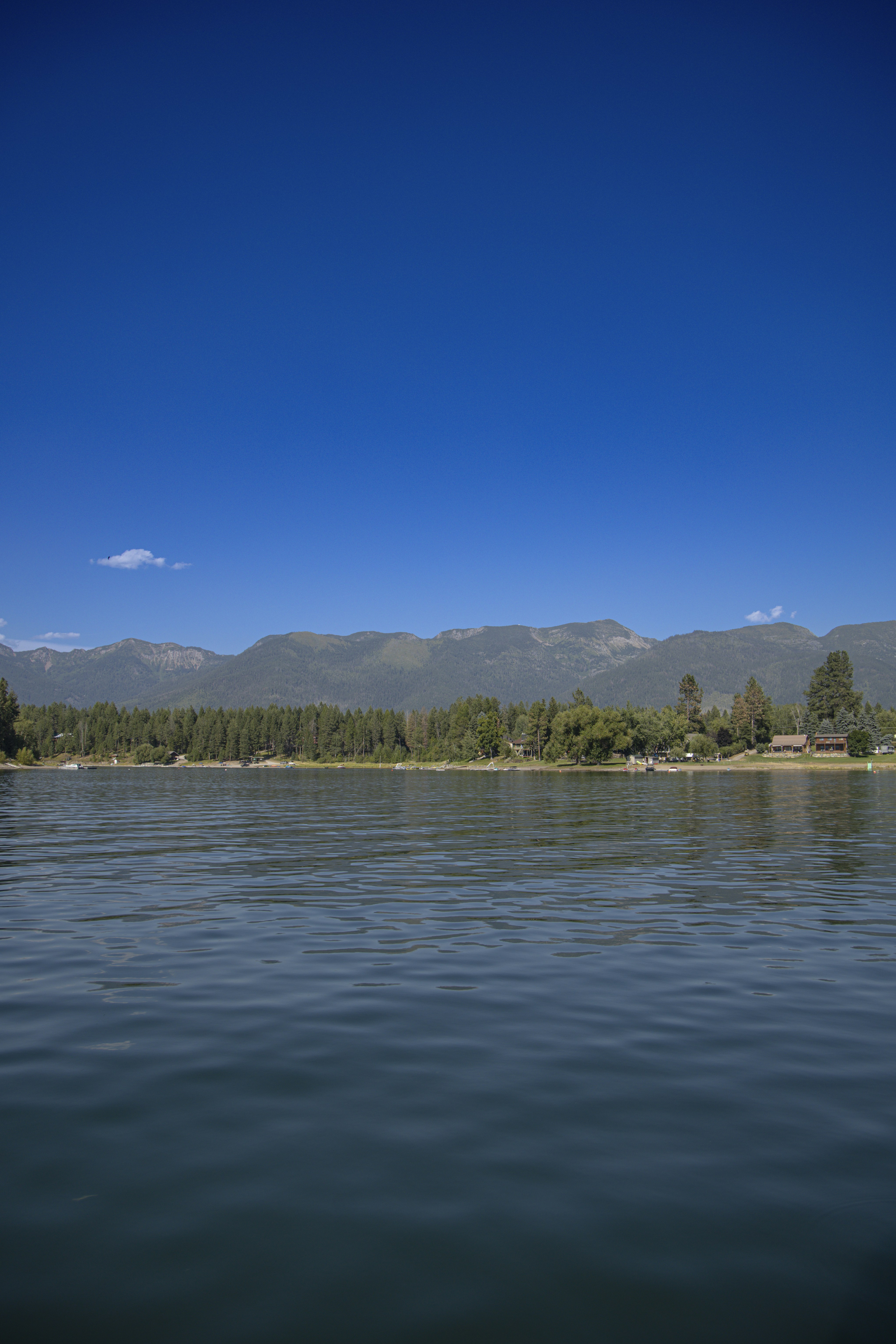 Calm lake reflecting the clear blue sky, bordered by lush greenery and distant mountains. The scene evokes a sense of peace and natural beauty.