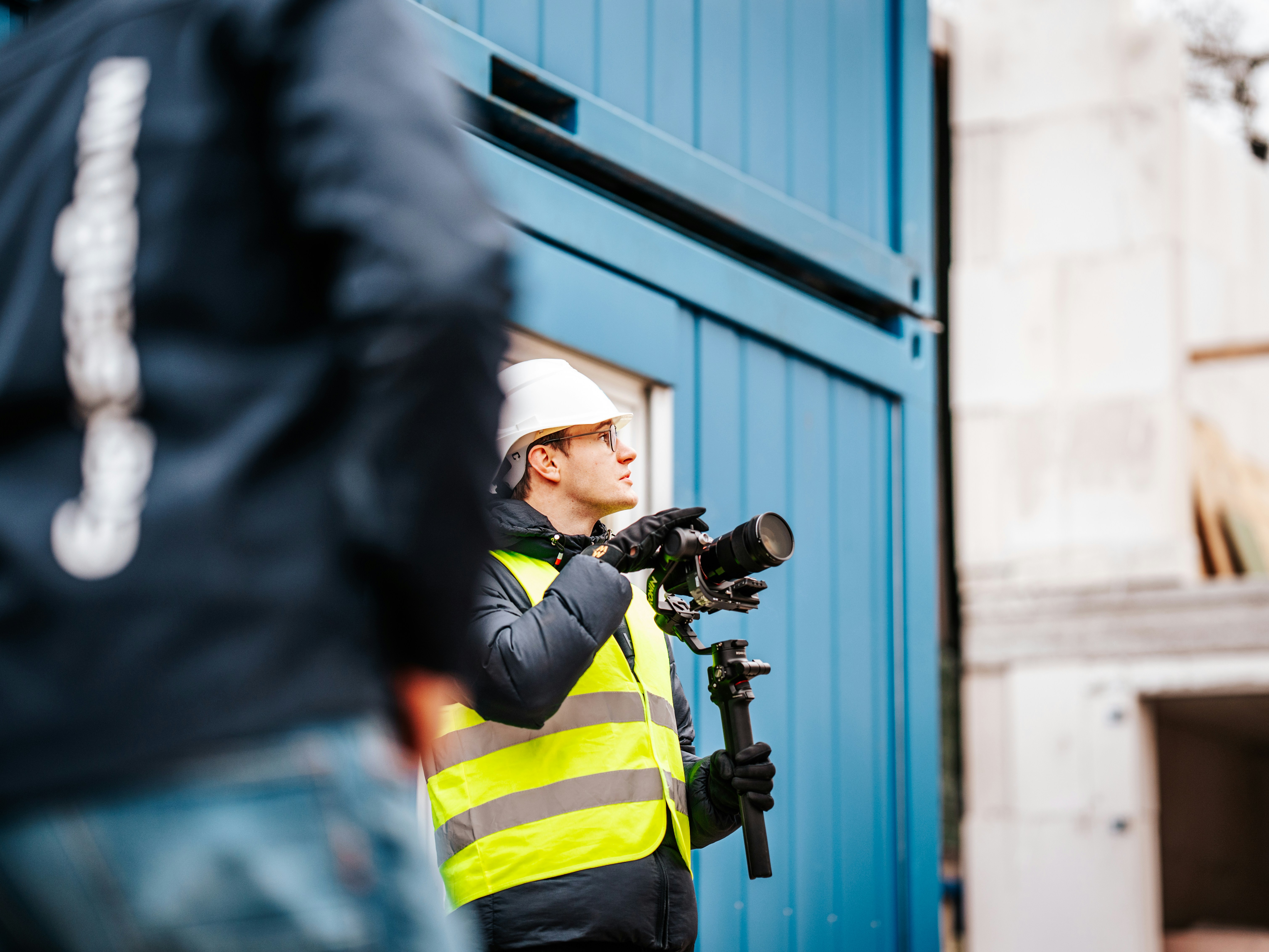 Stabilized Gaze: On-Site Photographer Bracing a Long Lens Against a Blue Construction Backdrop