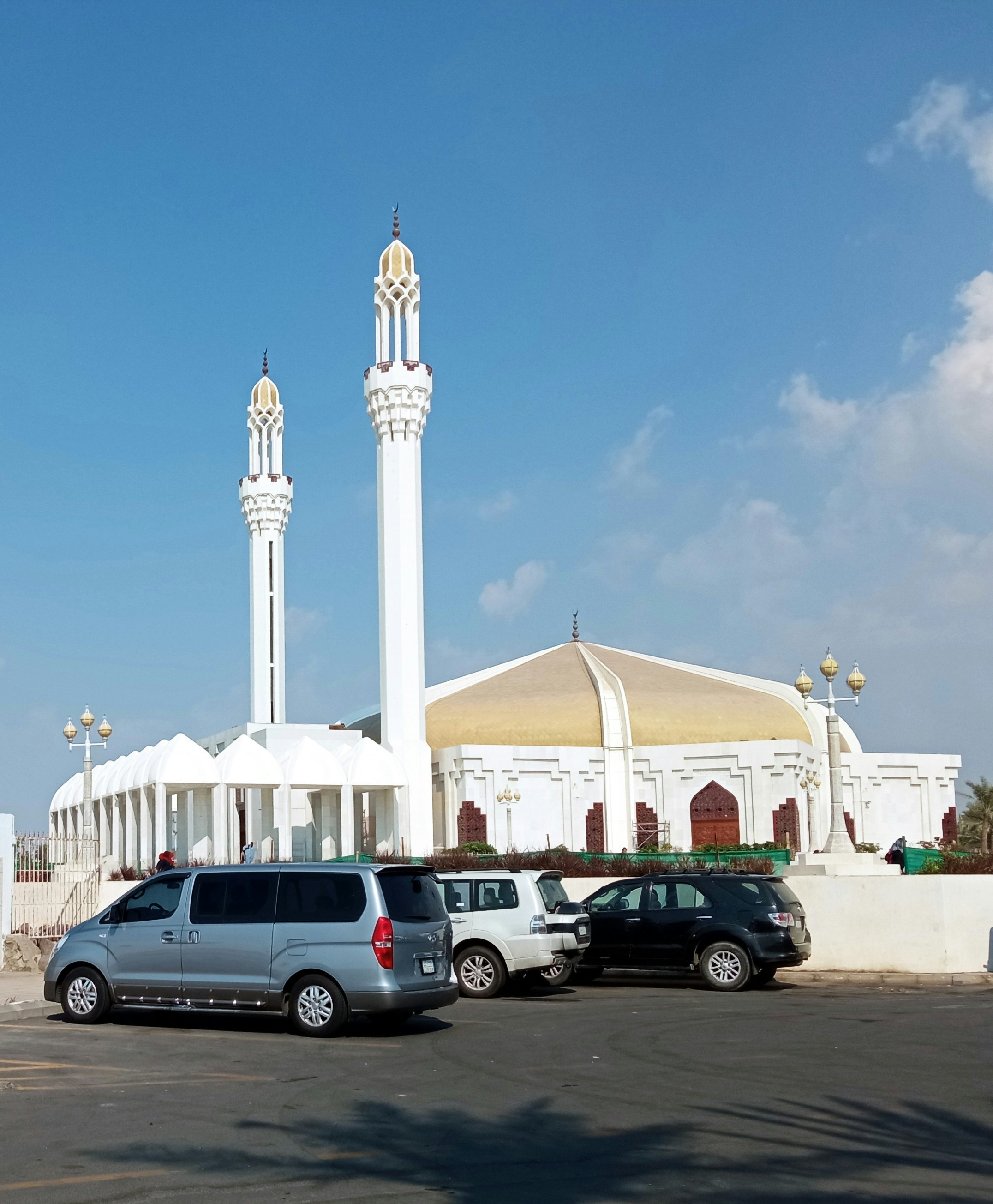 White mosque with two tall minarets set against a bright blue sky, with parked cars in the foreground.