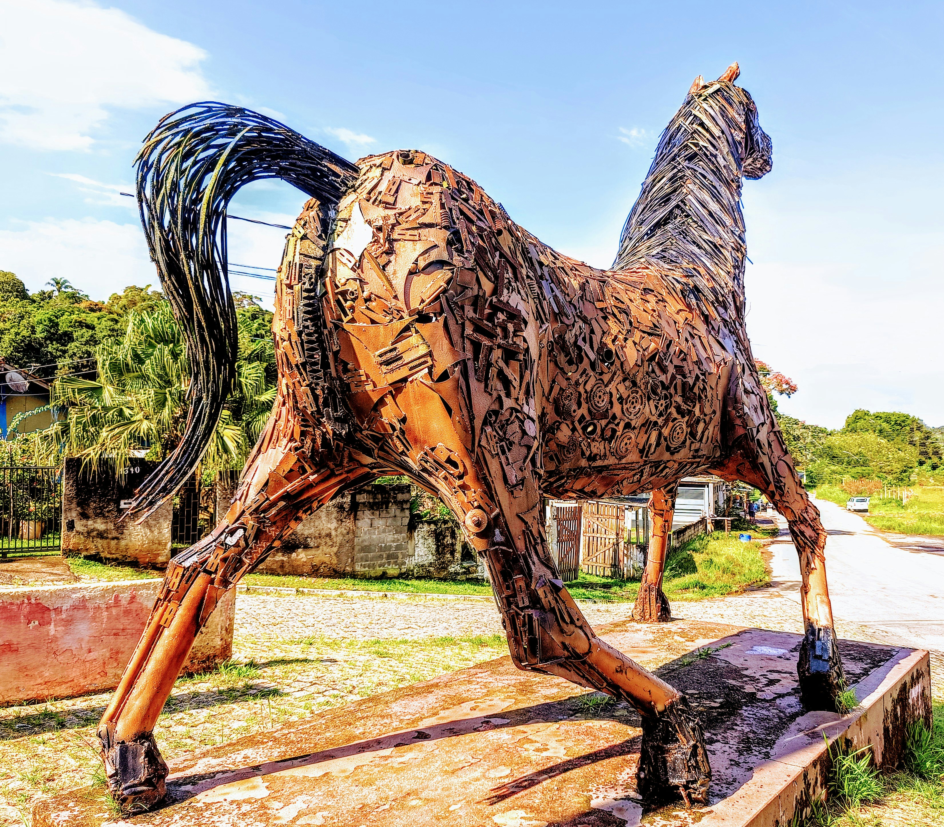 Metal scrap-metal horse sculpture stands in a sunlit park, with a clear blue sky and surrounding greenery.