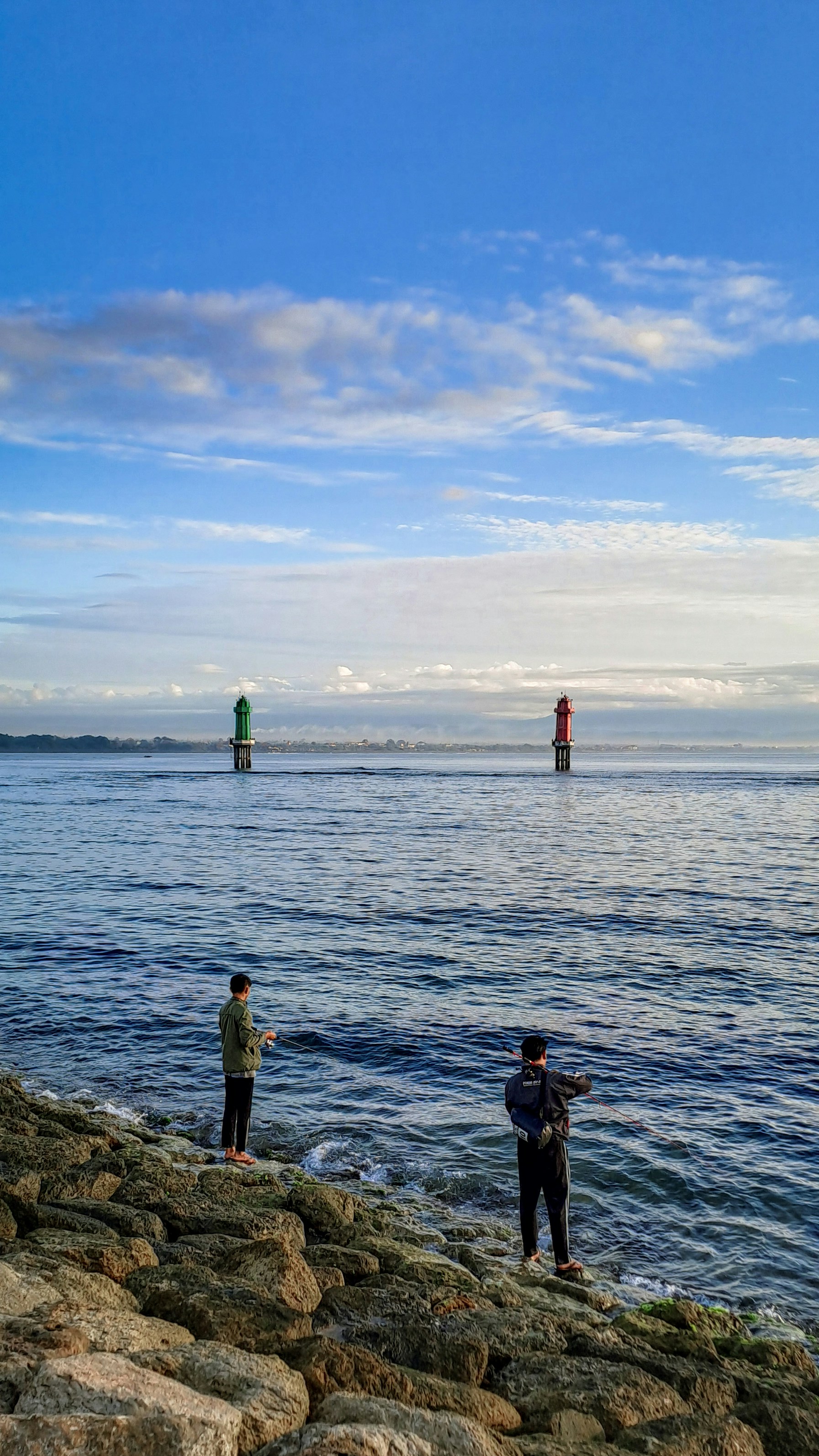 Two people fishing on a rocky shore with green and red beacons in the distance under a clear blue sky.