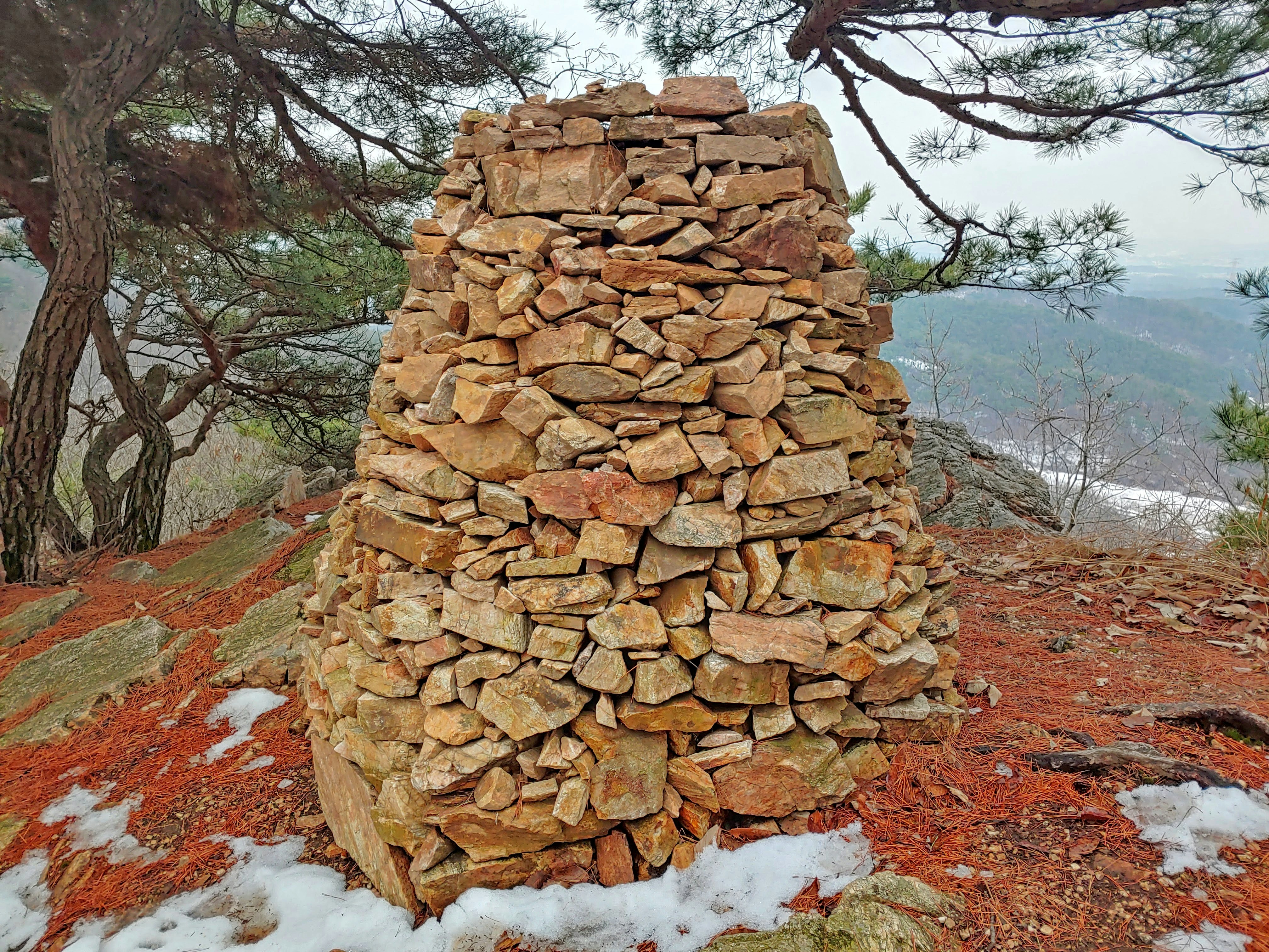 A rugged stone cairn rises on a red-earth ridge, with snow patches and pine trees framing distant hills.