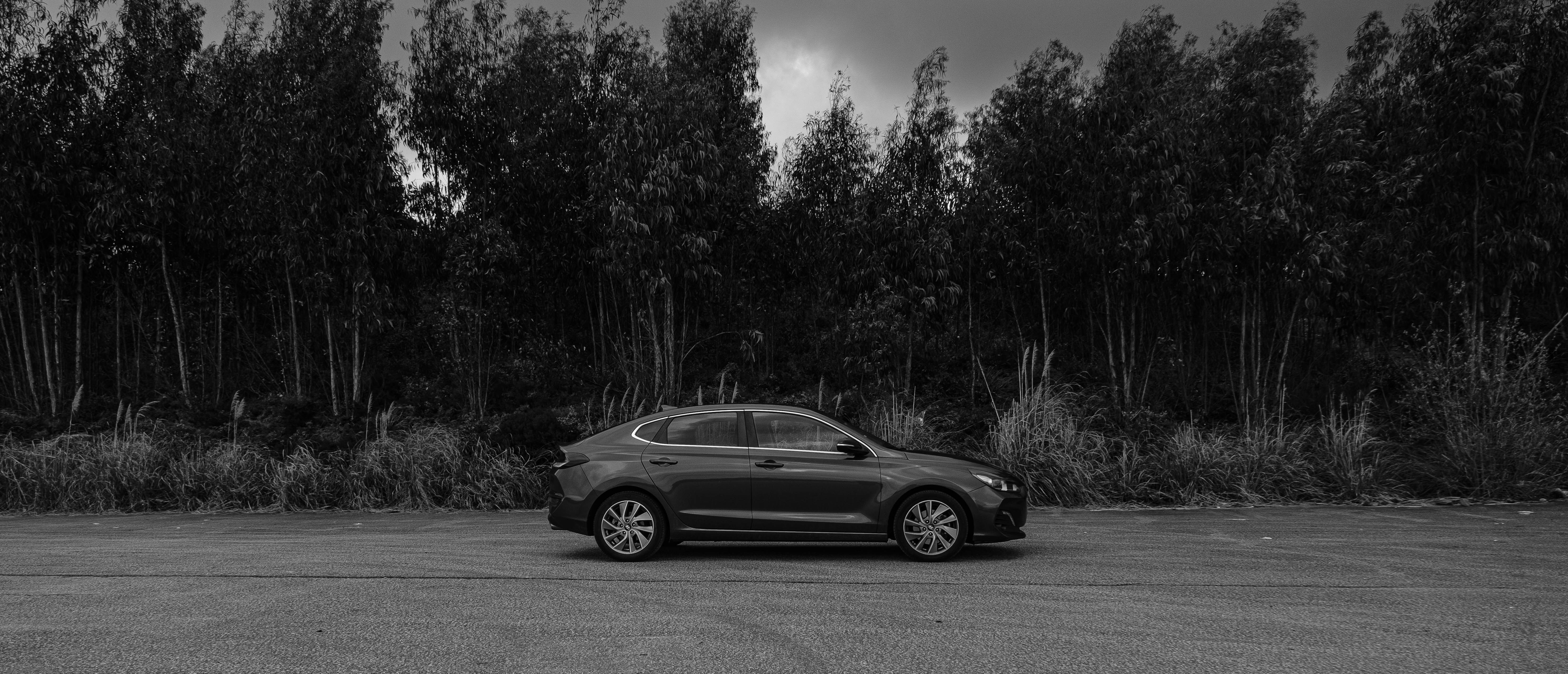 Dark sedan parked on a dirt road with a dense forest backdrop under a cloudy sky.