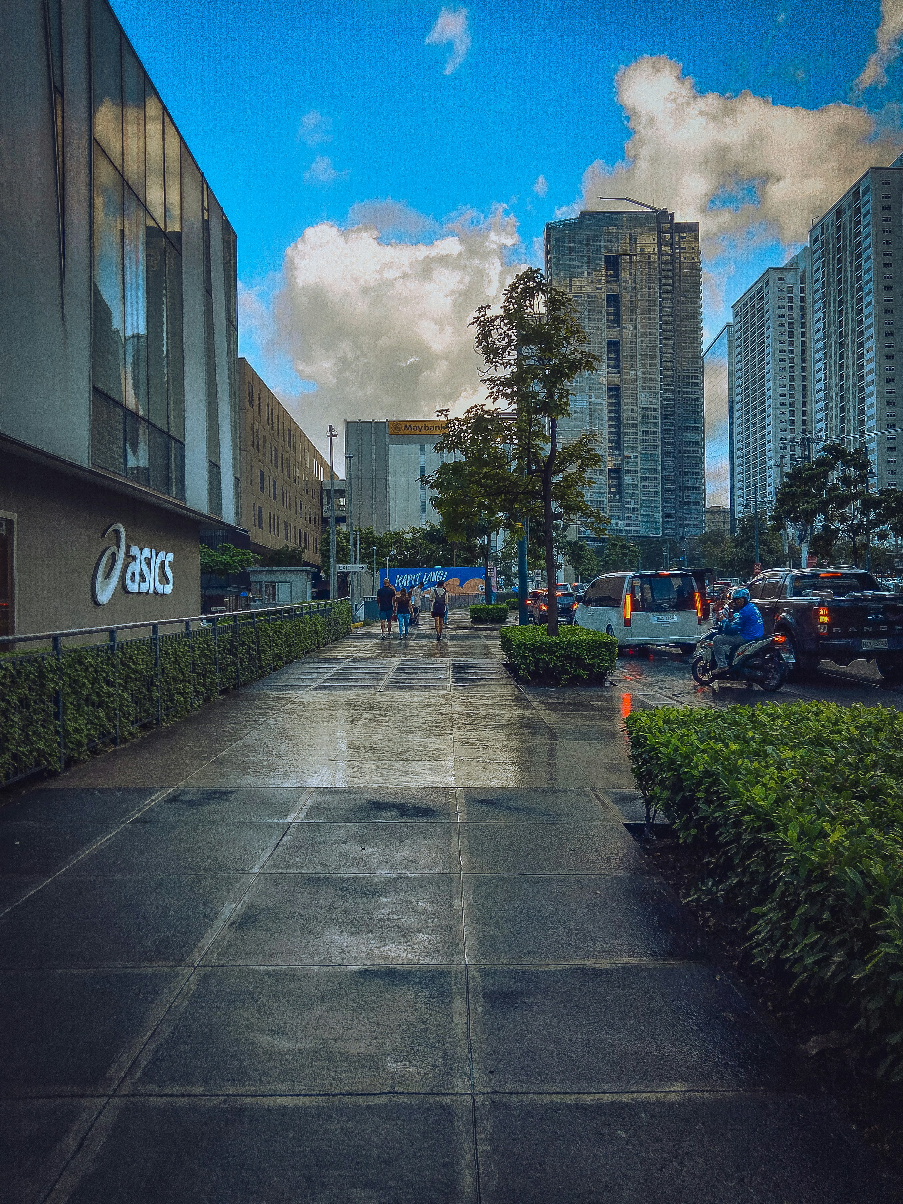 Wet city sidewalk reflects light after rain as pedestrians move toward the skyline. The left foreground features a storefront with the Asics logo.