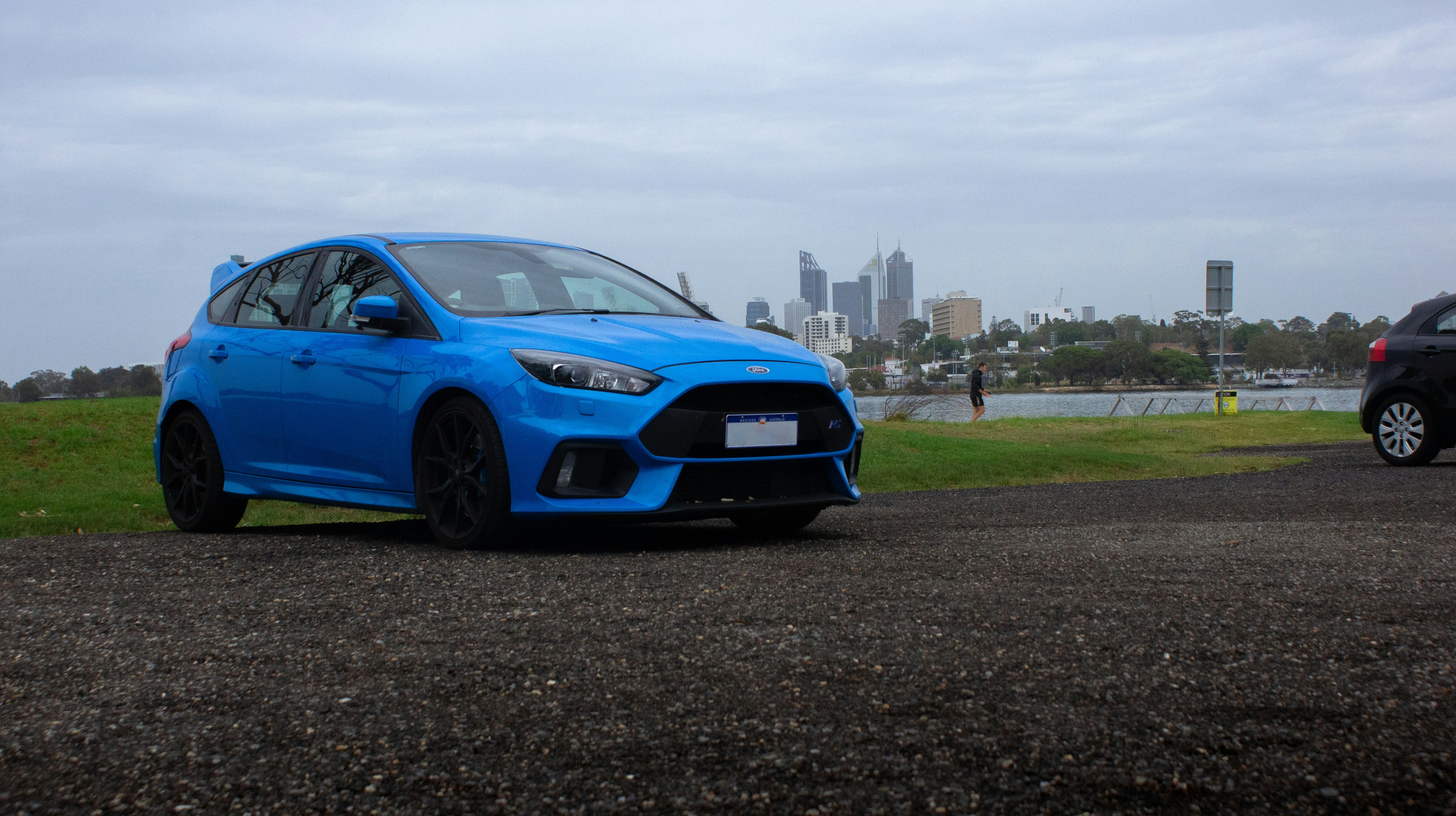 Azure Streak: a blue hatchback rests beside a lakeside road with a distant city skyline