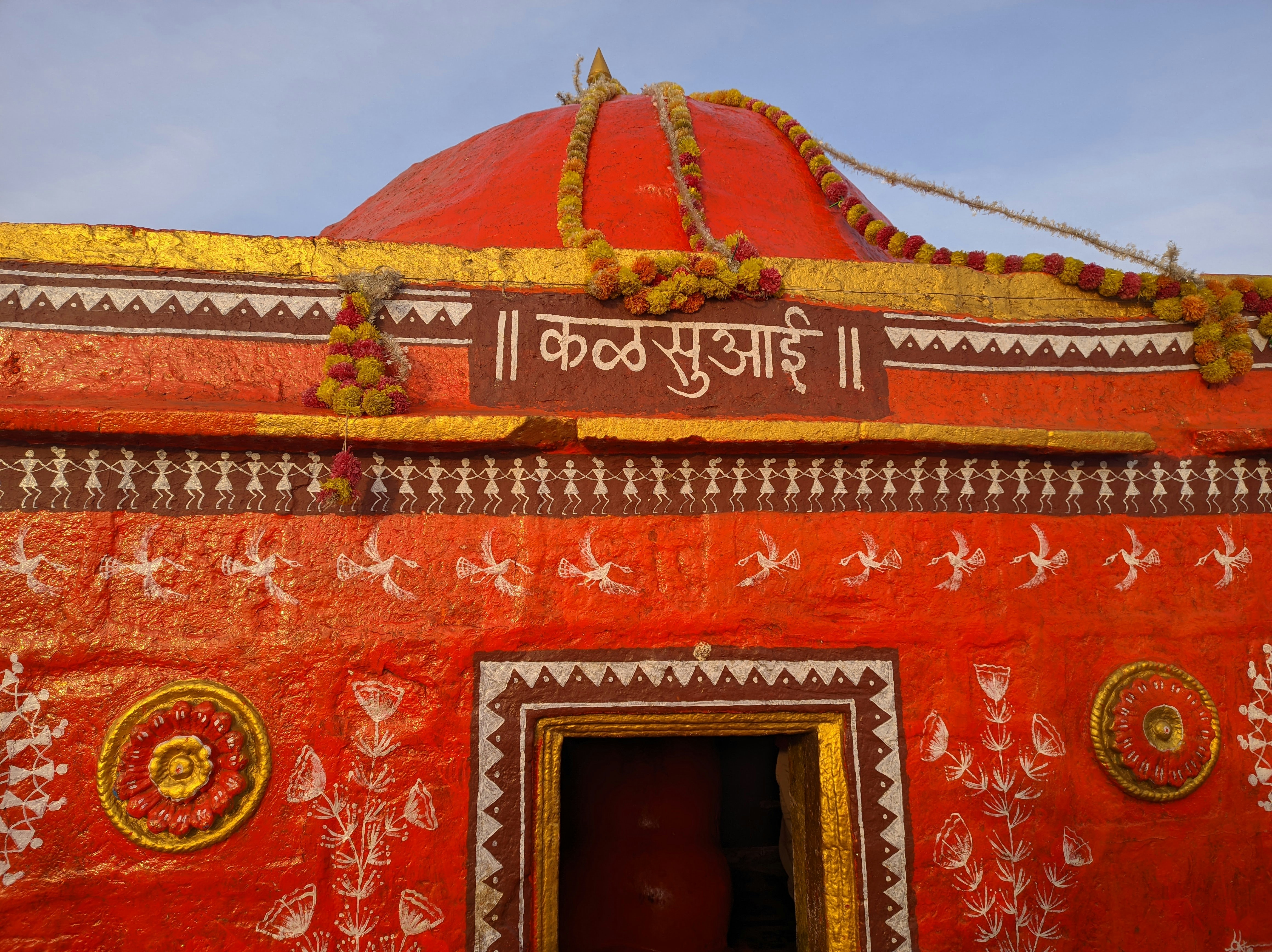 Crimson temple facade adorned with marigold garlands and white geometric motifs centers on a dark doorway. Sunlit exterior reveals vivid textures and decorative inscriptions.