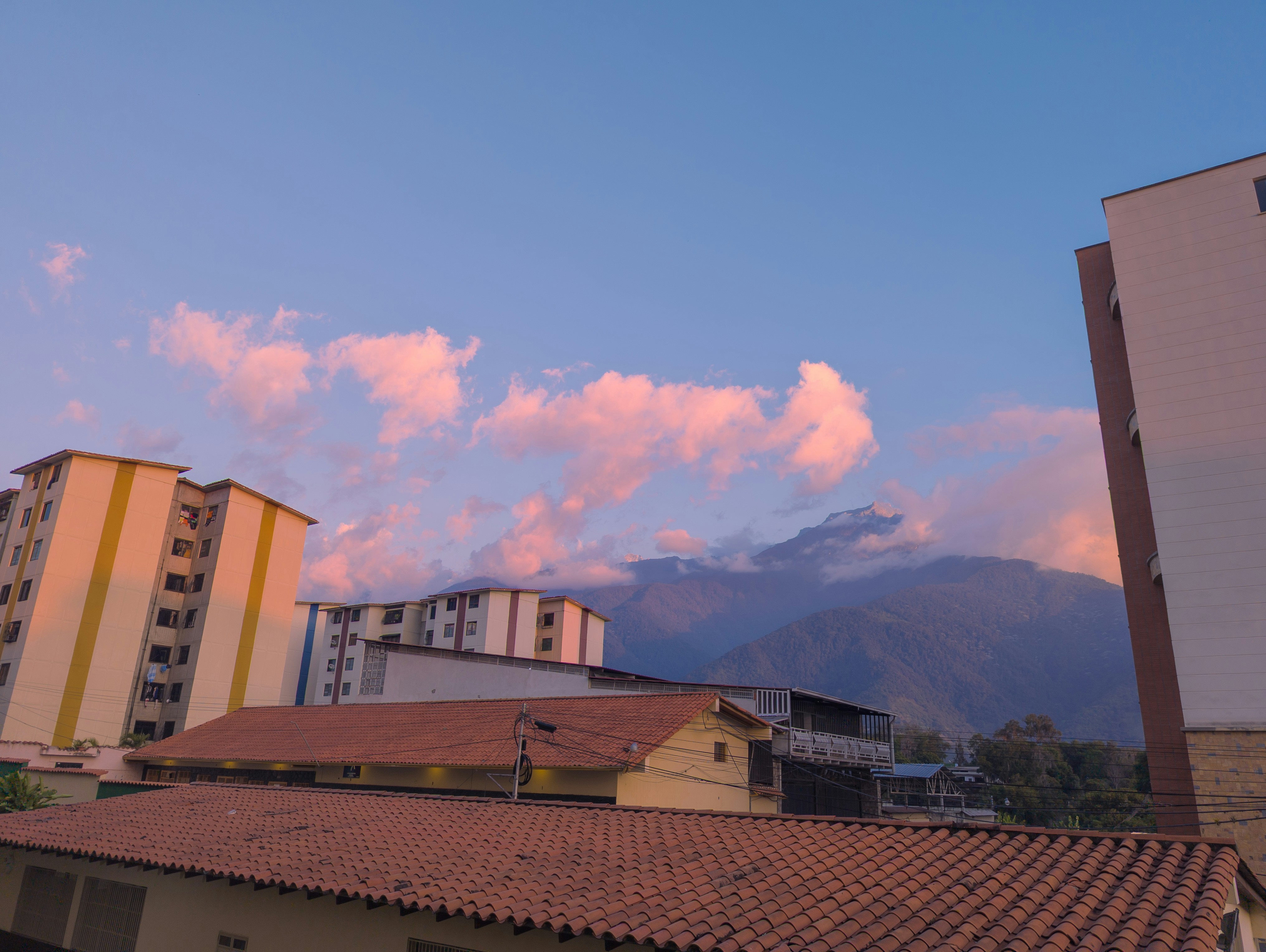 Soft pink clouds drift above urban buildings, with majestic mountains in the background. The scene captures the serene transition from night to day.