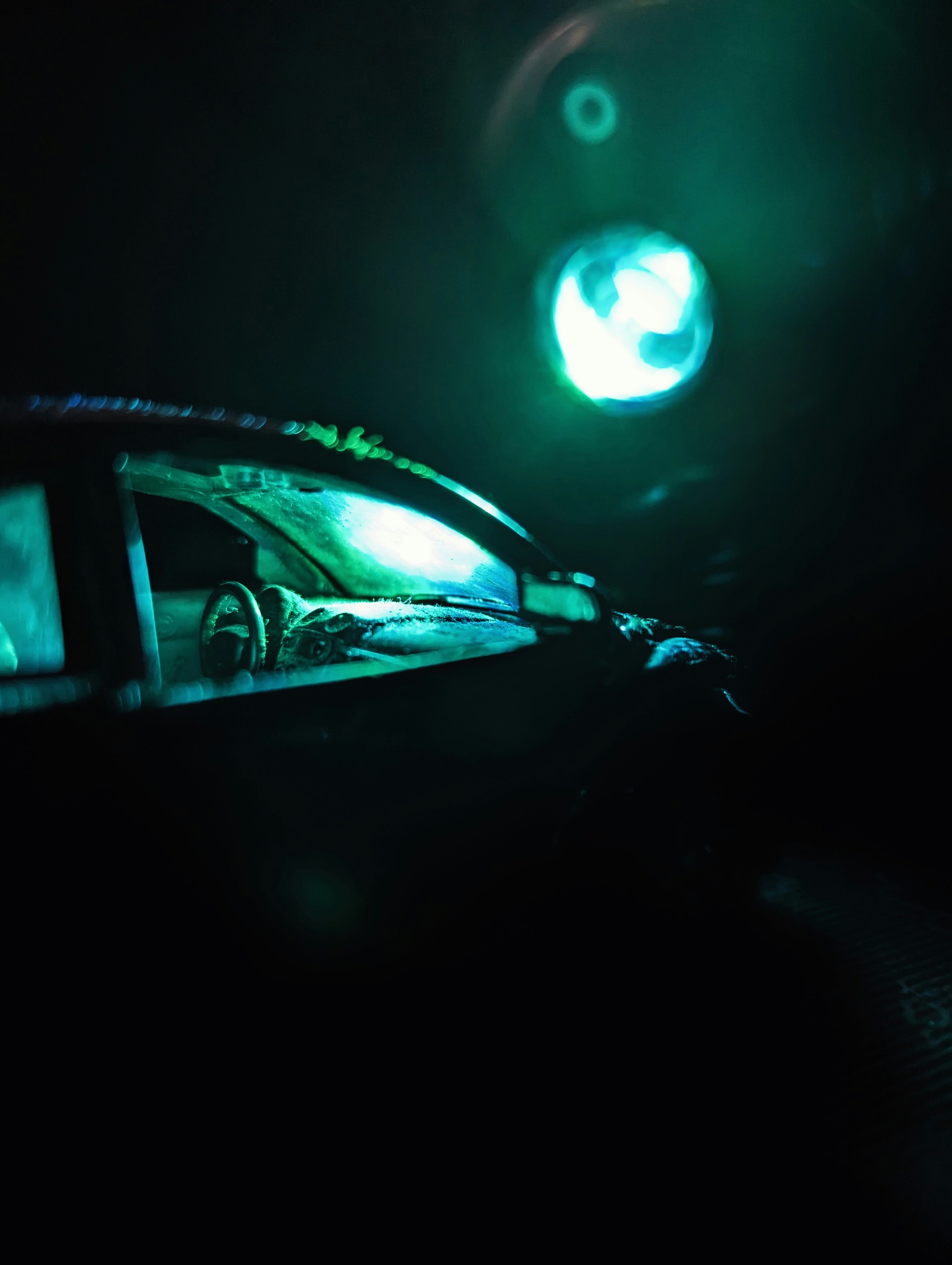 Teal-tinged night photograph of a weathered car interior, with a bright teal glow outside the window casting dramatic light on the dash.