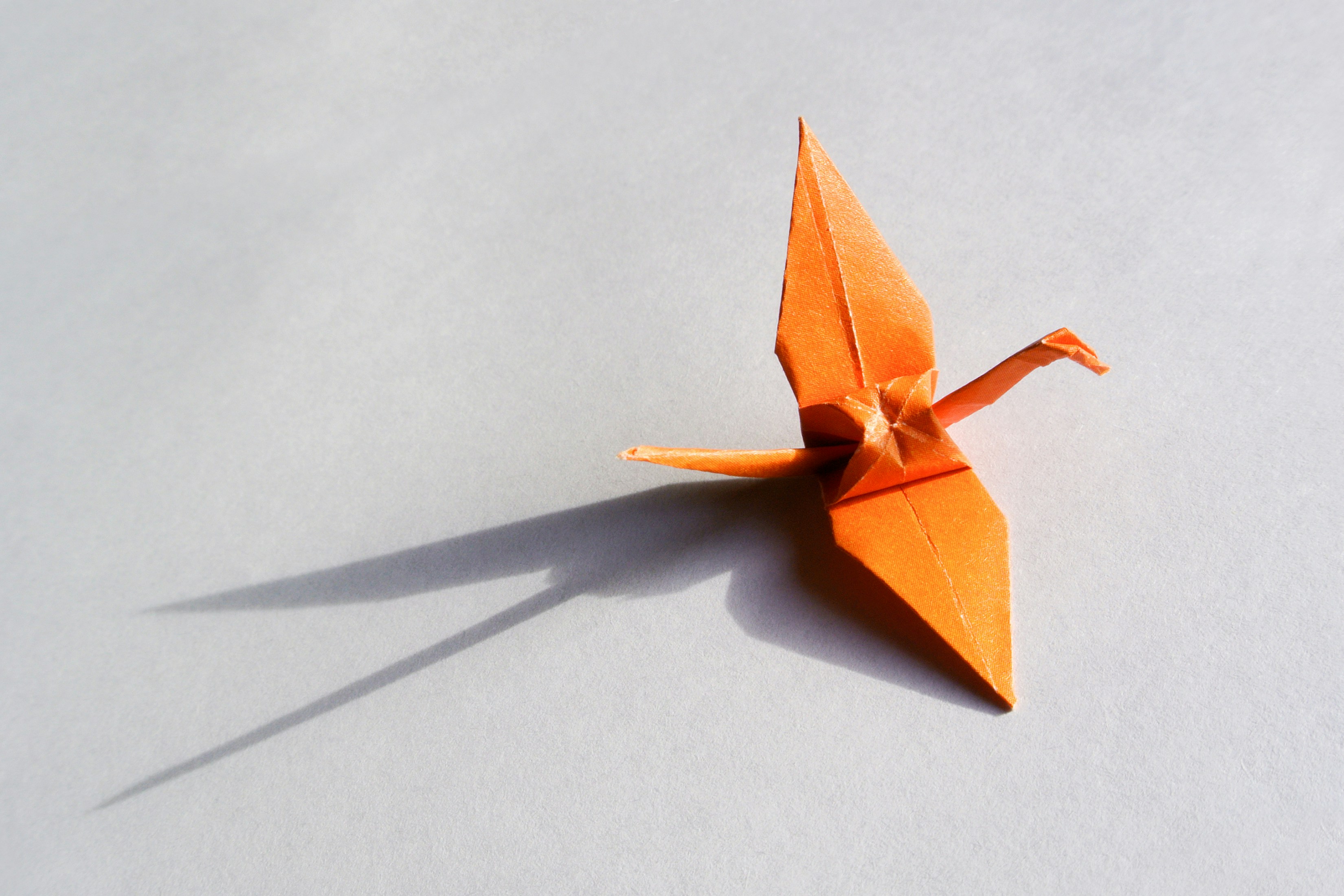 An orange origami crane delicately poised on a light gray surface, casting a subtle shadow that enhances its three-dimensional form.