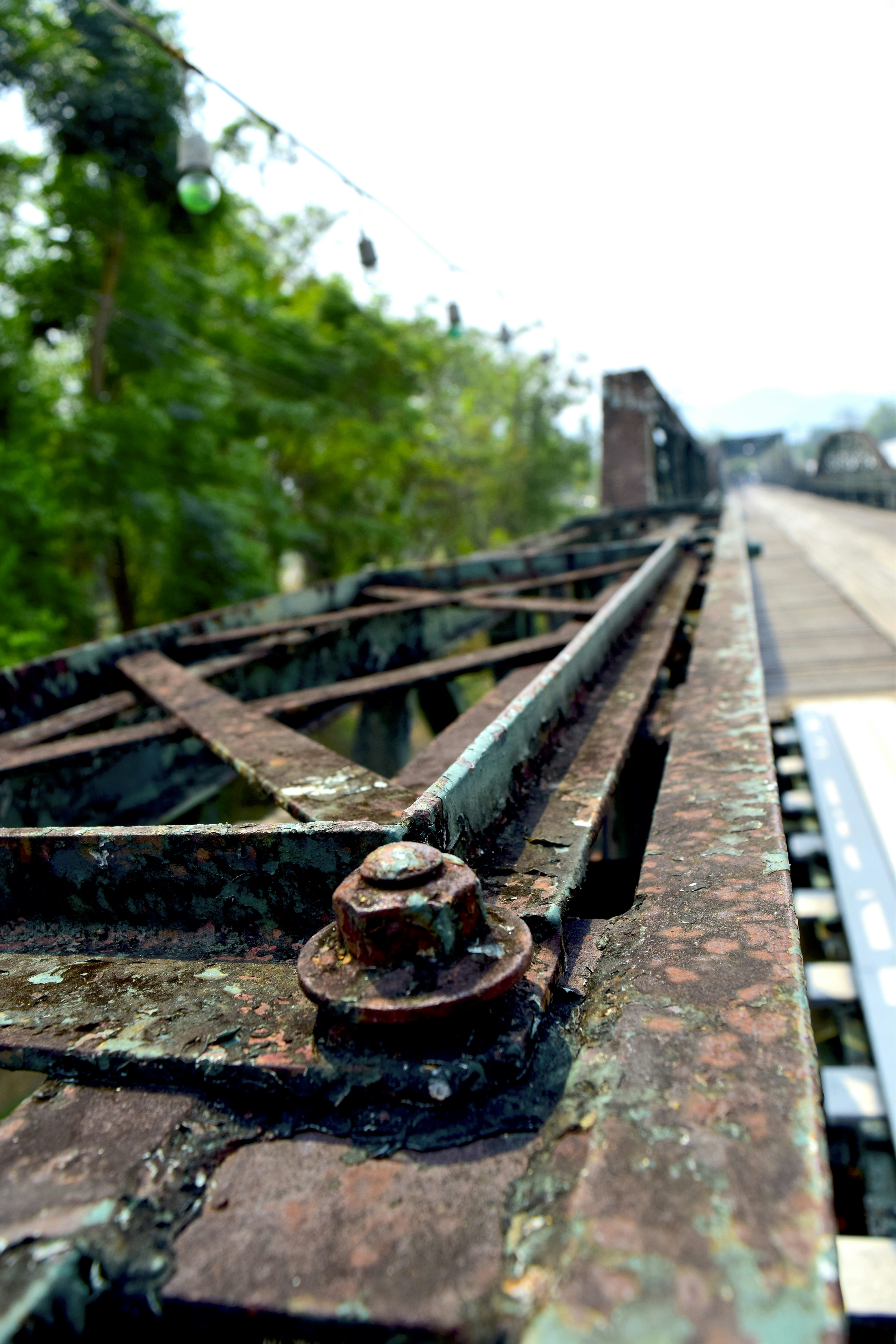 Close-up view of a weathered metal beam on an old bridge, showcasing rust and decay against a backdrop of lush greenery.