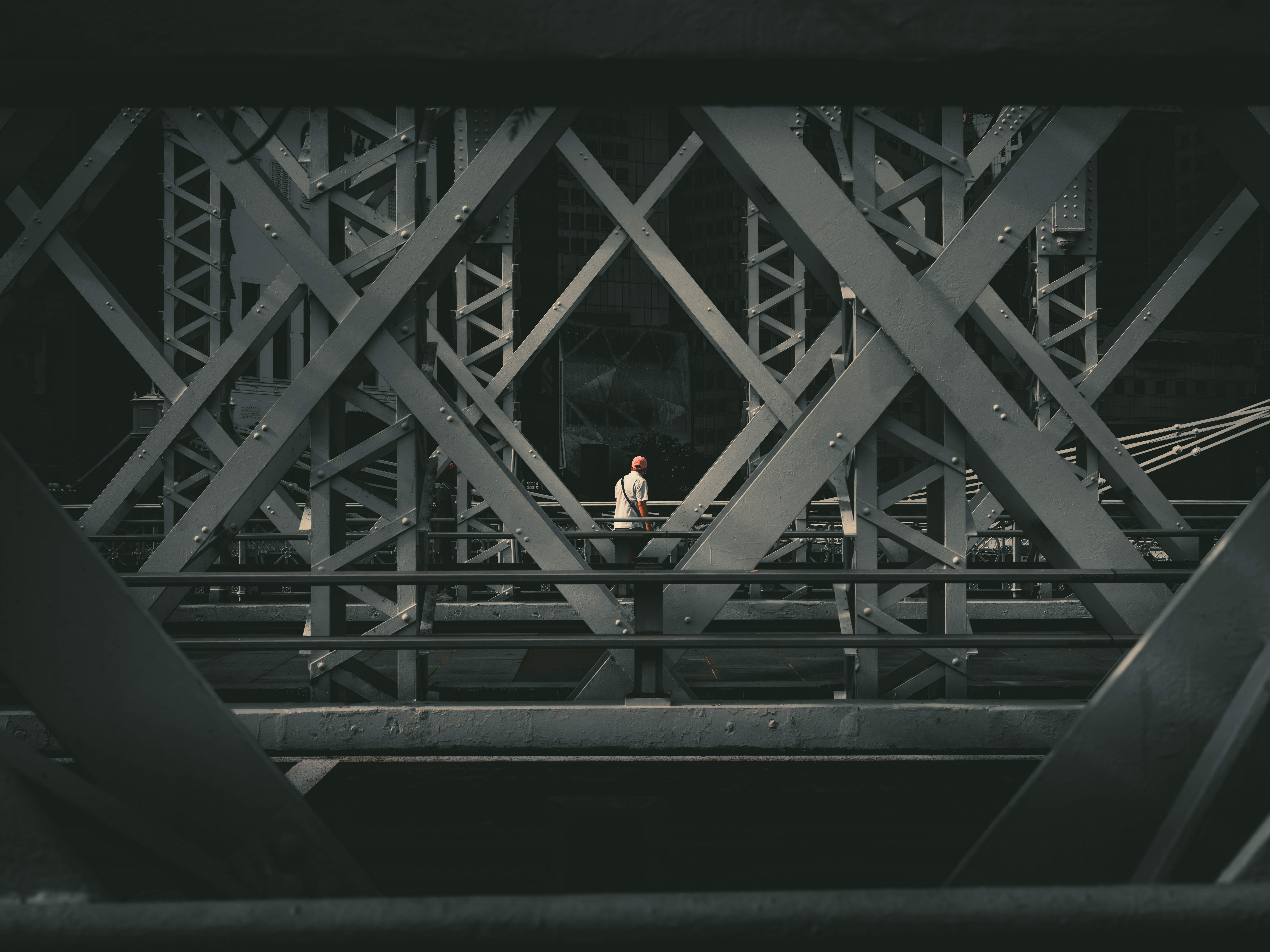 A lone man in an orange shirt stands amid a crisscrossed steel bridge framework.
