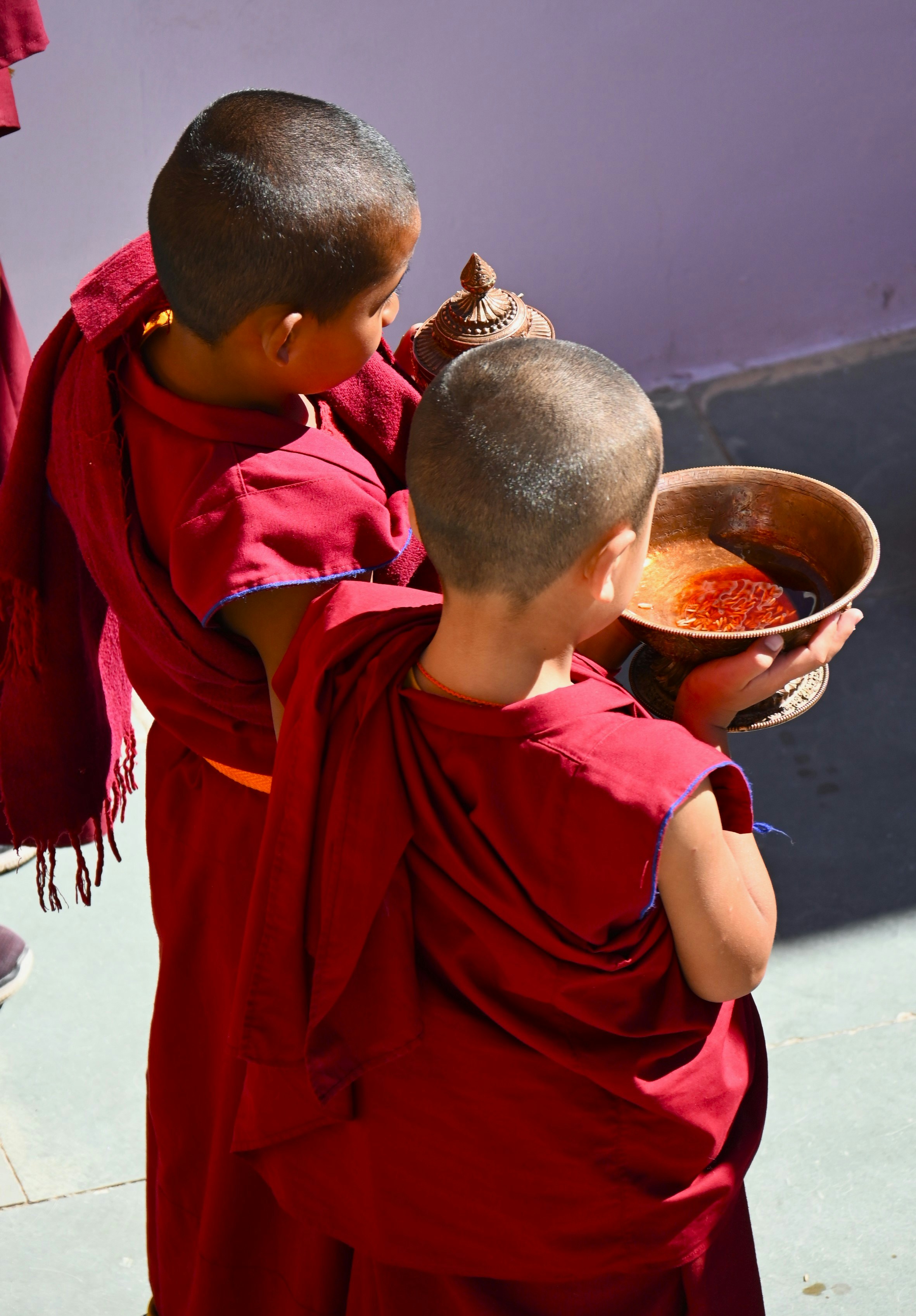 Crimson-robed novices cradle a copper alms bowl in bright courtyard light