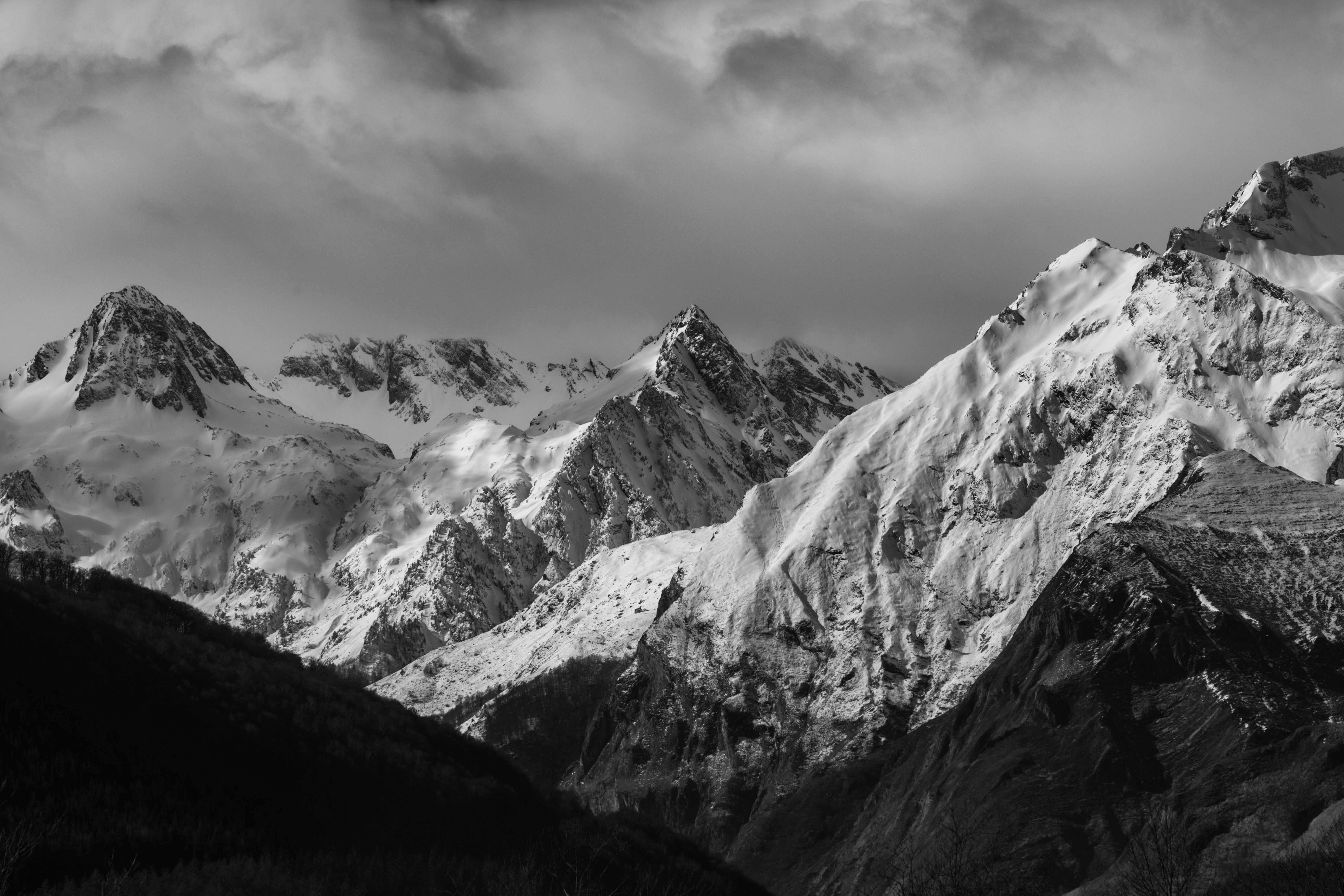 Snow-capped mountain range under dramatic clouds, captured in black and white. The rugged terrain showcases the raw beauty of nature.