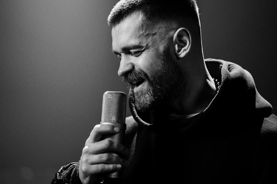 A man sings into a microphone in a dramatic black-and-white portrait, lit from above and the side. He wears a dark hoodie and smiles with eyes closed.