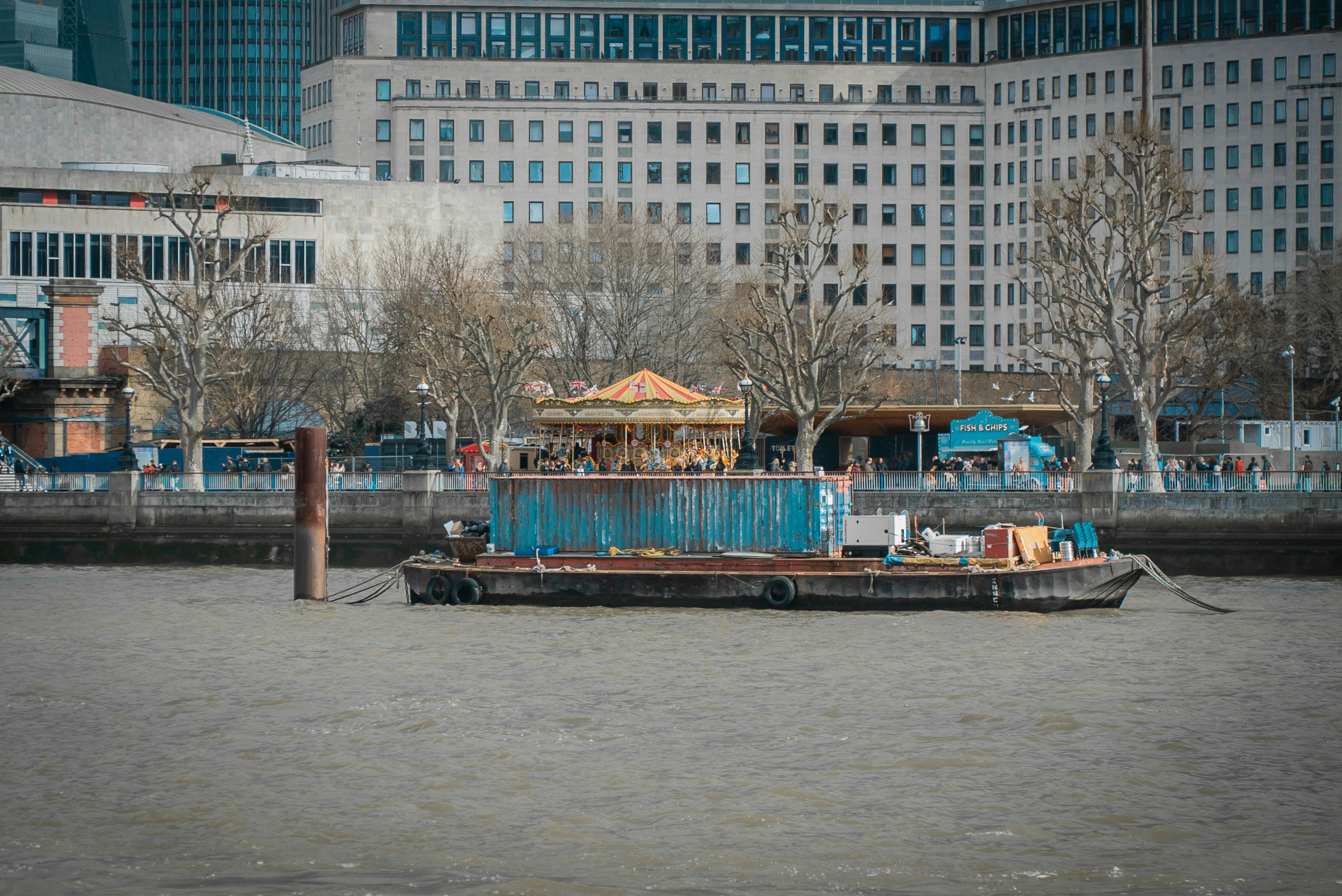Blue cargo barge with orange canopy gliding along a city riverfront