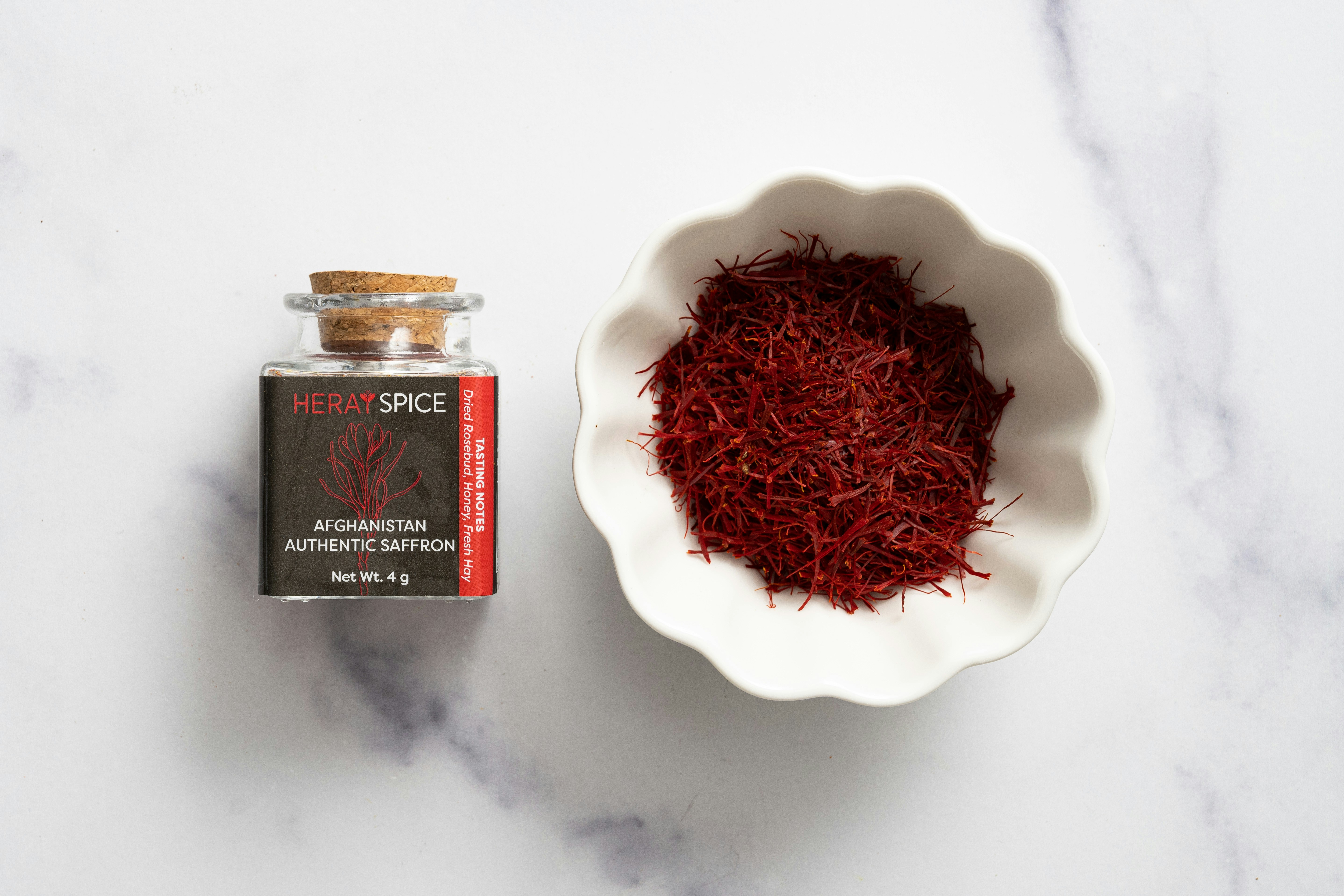 White bowl filled with red saffron threads next to a glass jar of spice on a marble surface.