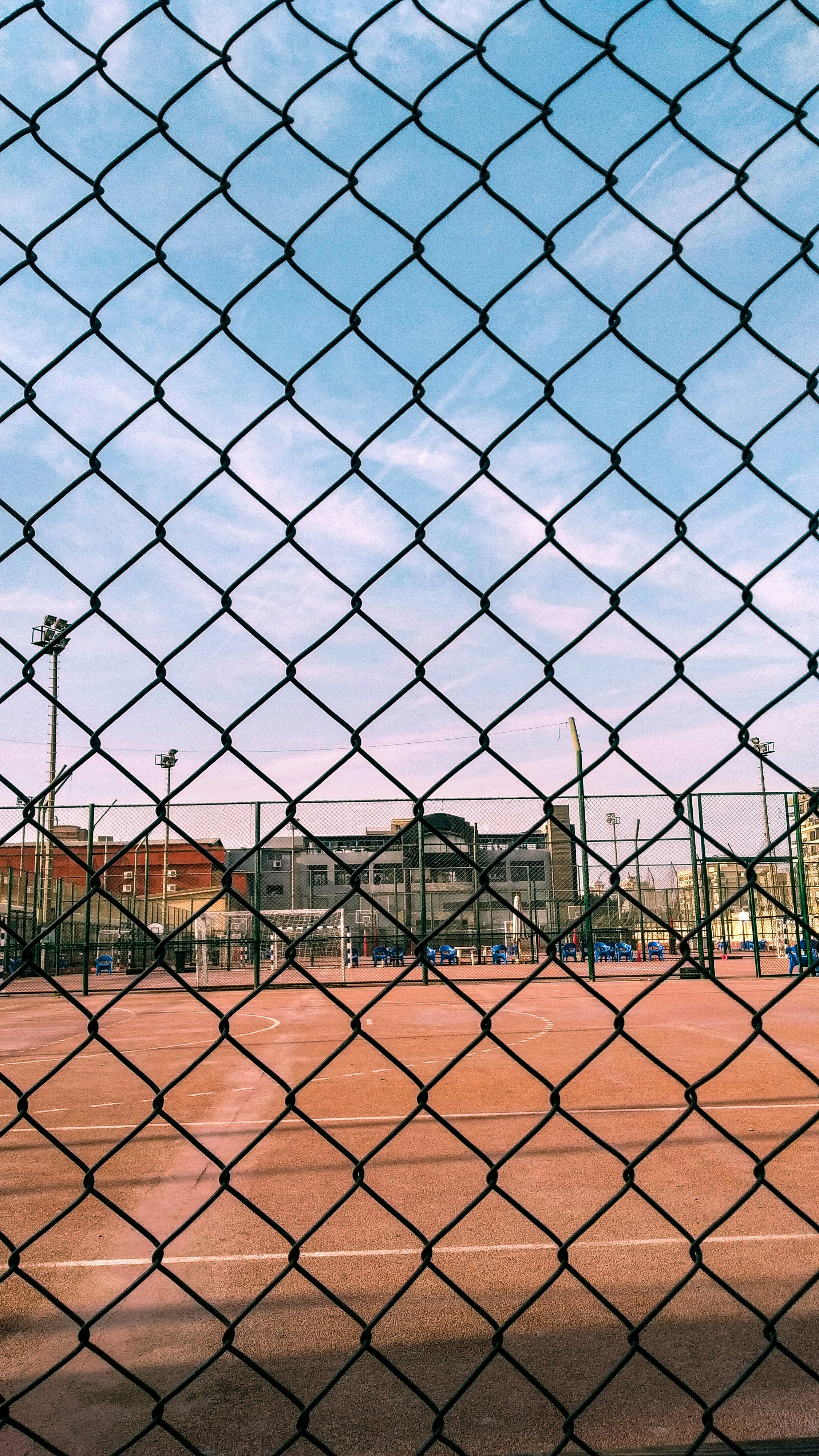 Chain-link mesh fills the foreground, opening to a clay sports field and distant buildings beneath a pastel sky.