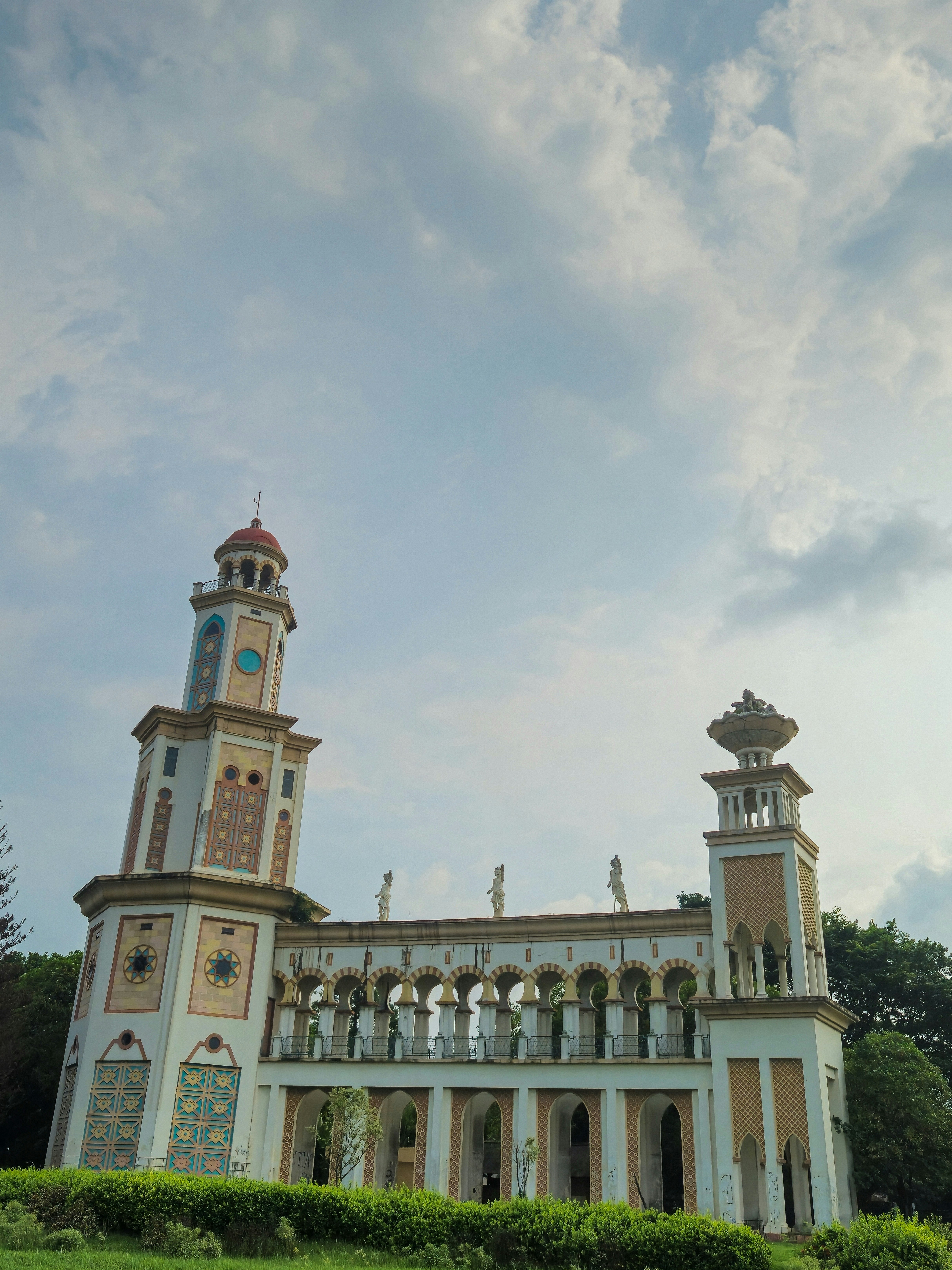 A photograph of a colorful ornamental courtyard pavilion featuring a red-domed tower and an arched colonnade beneath a bright sky.