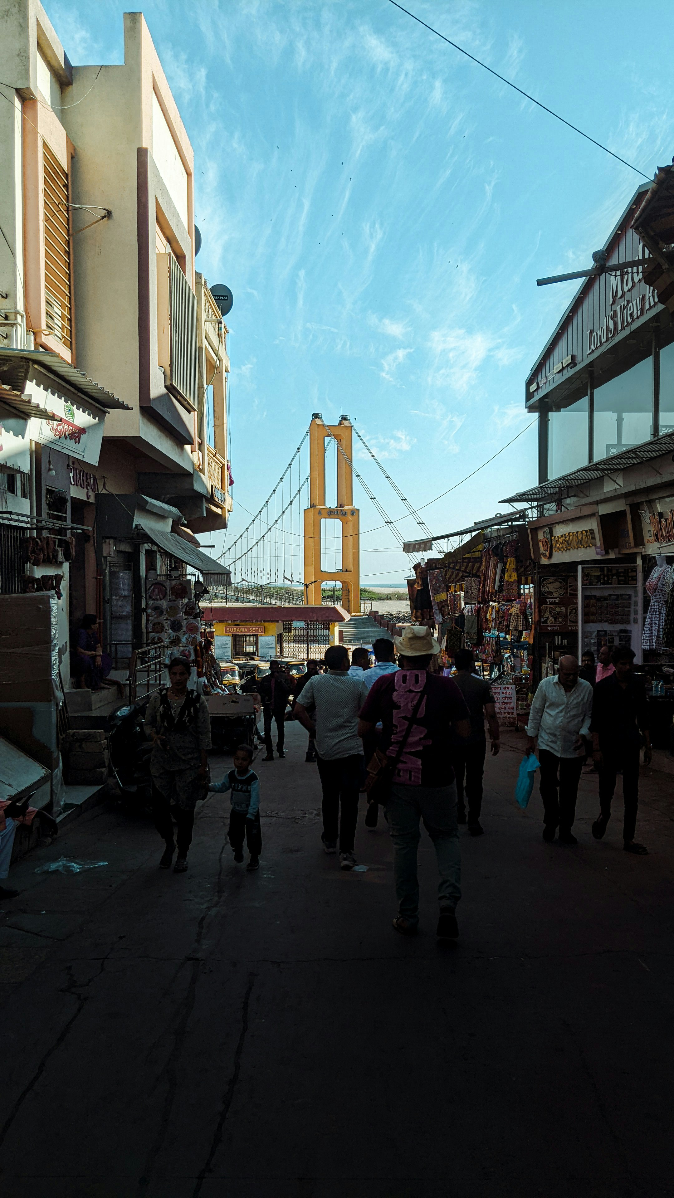 People walking through a vibrant market street towards the Sudhama Setu bridge under a clear sky.
