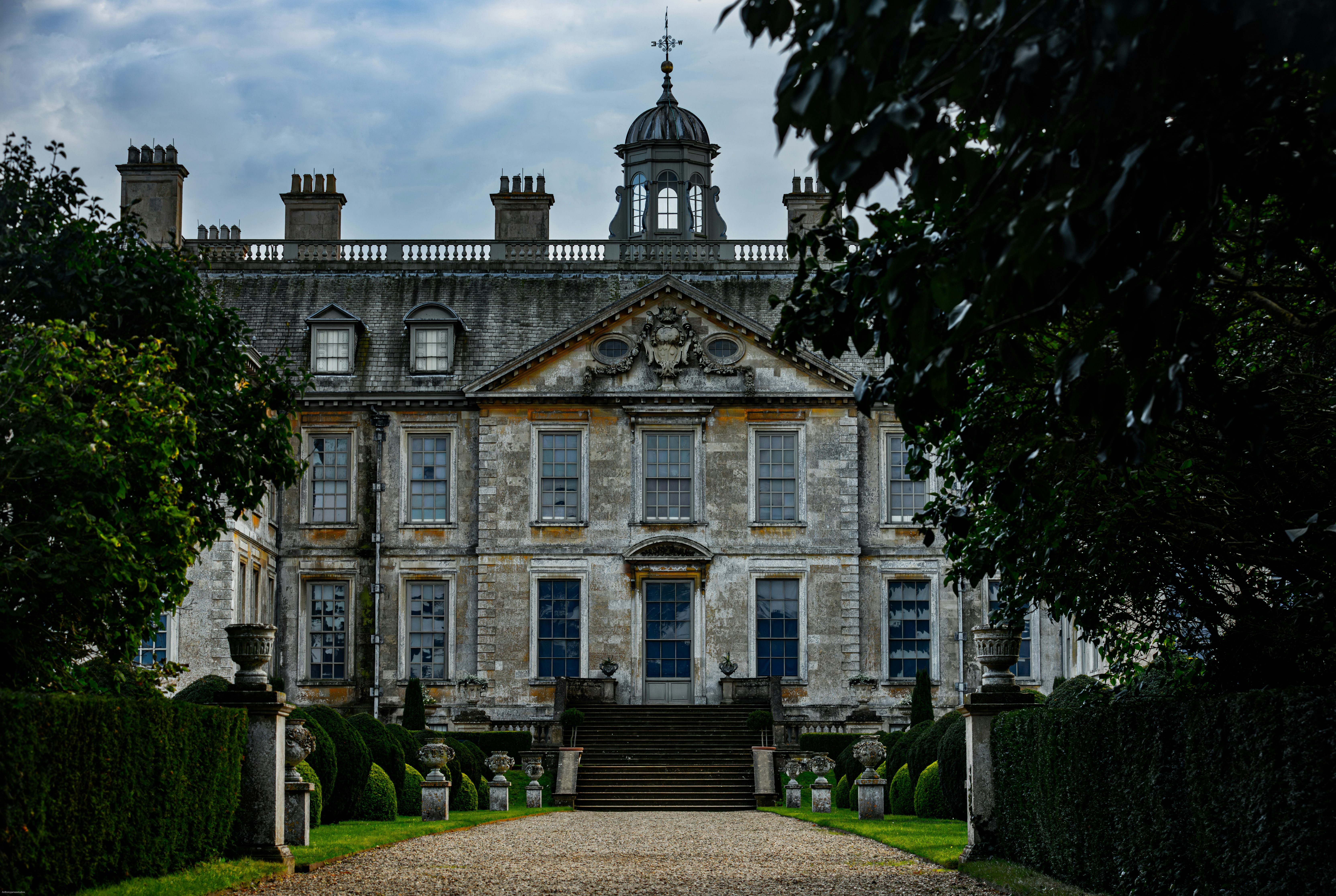 Grand historic mansion framed by lush green trees under a cloudy sky.