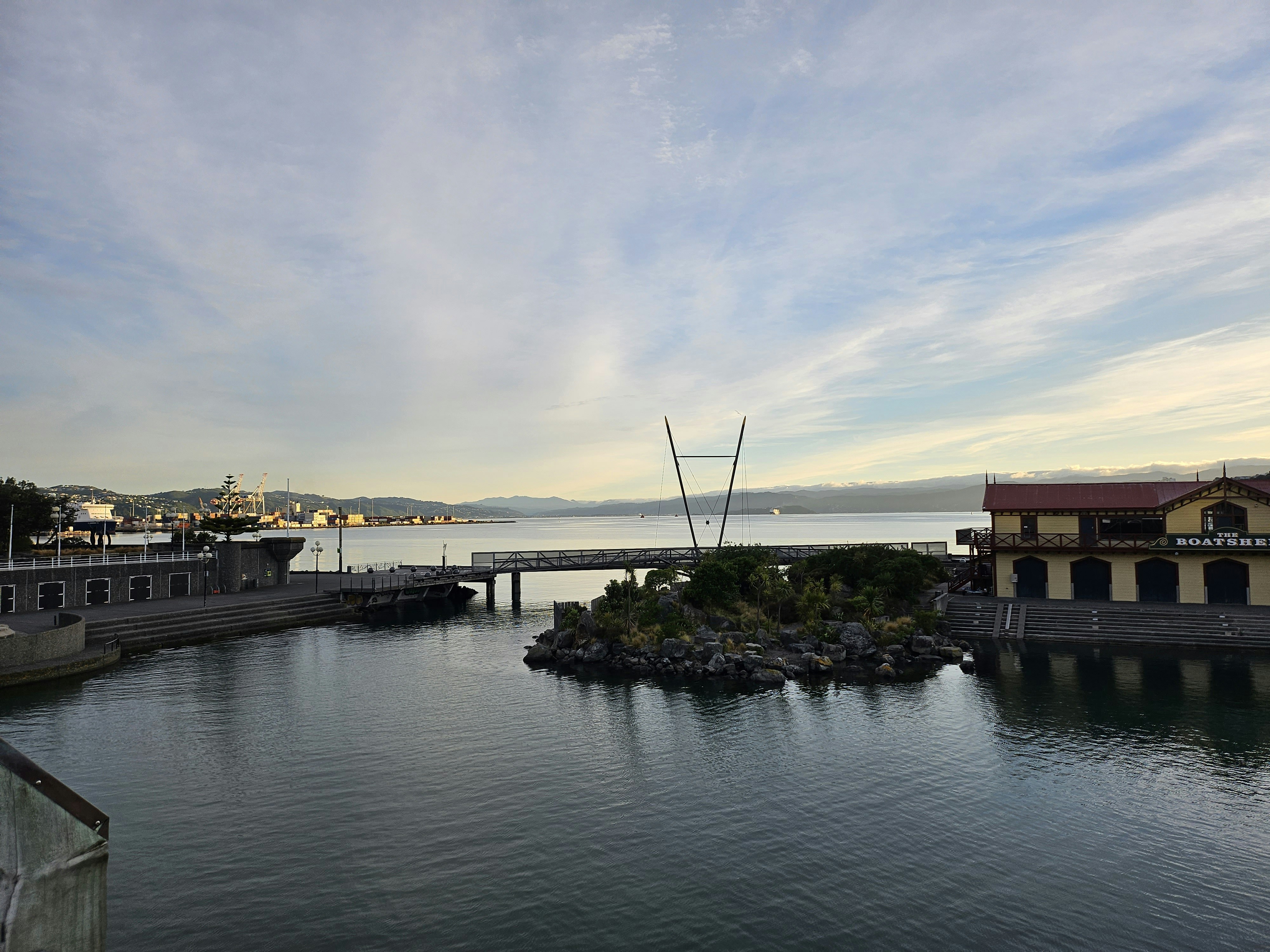 A tranquil harbor scene featuring a small island and a rustic building along the water's edge. The calm surface reflects the sky and surrounding structures.