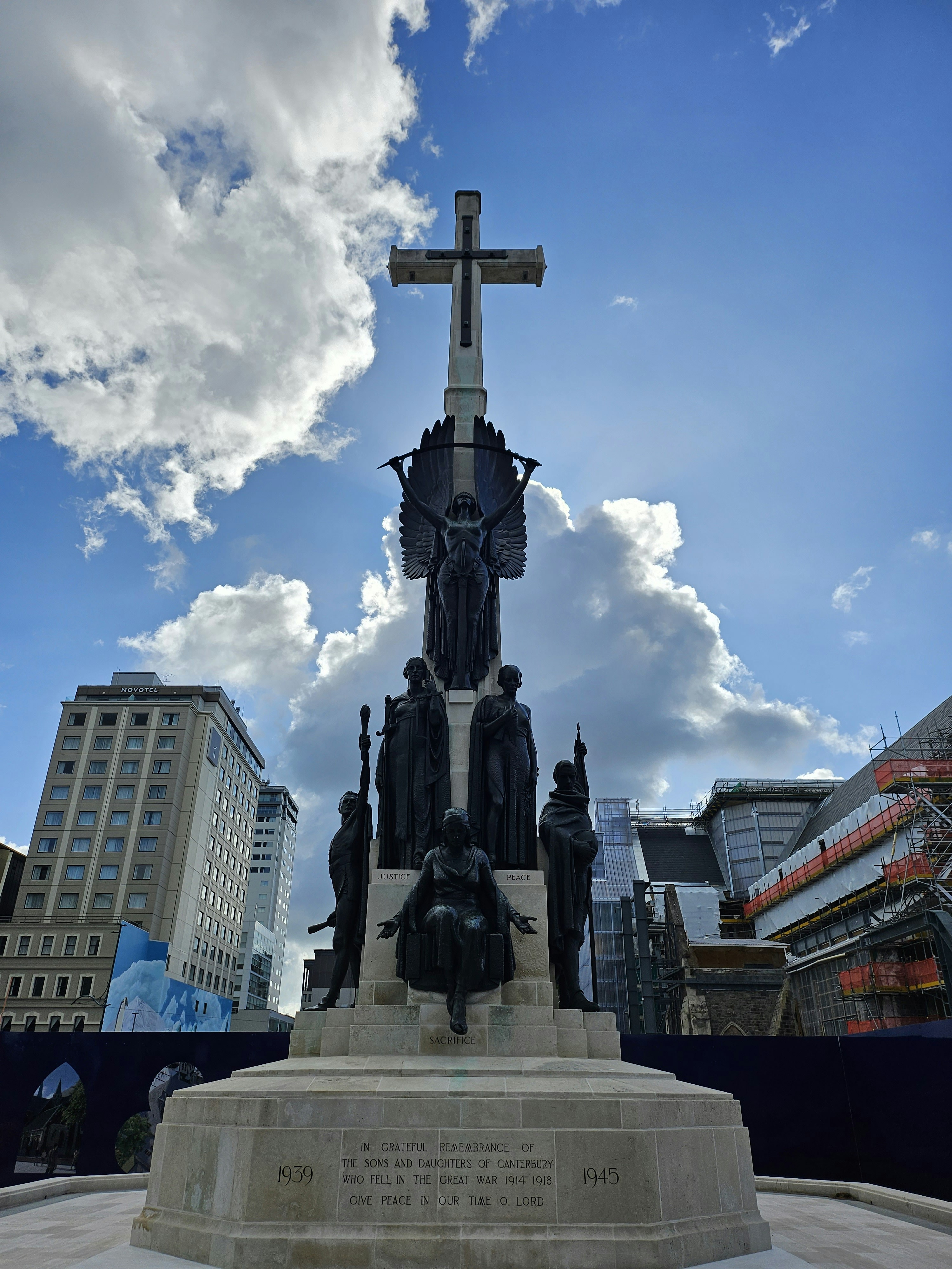 A tall bronze monument with a cross at the summit rises above life-sized statues gathered on a stepped pedestal. The scene is set in a modern city square under a blue sky with fluffy clouds.