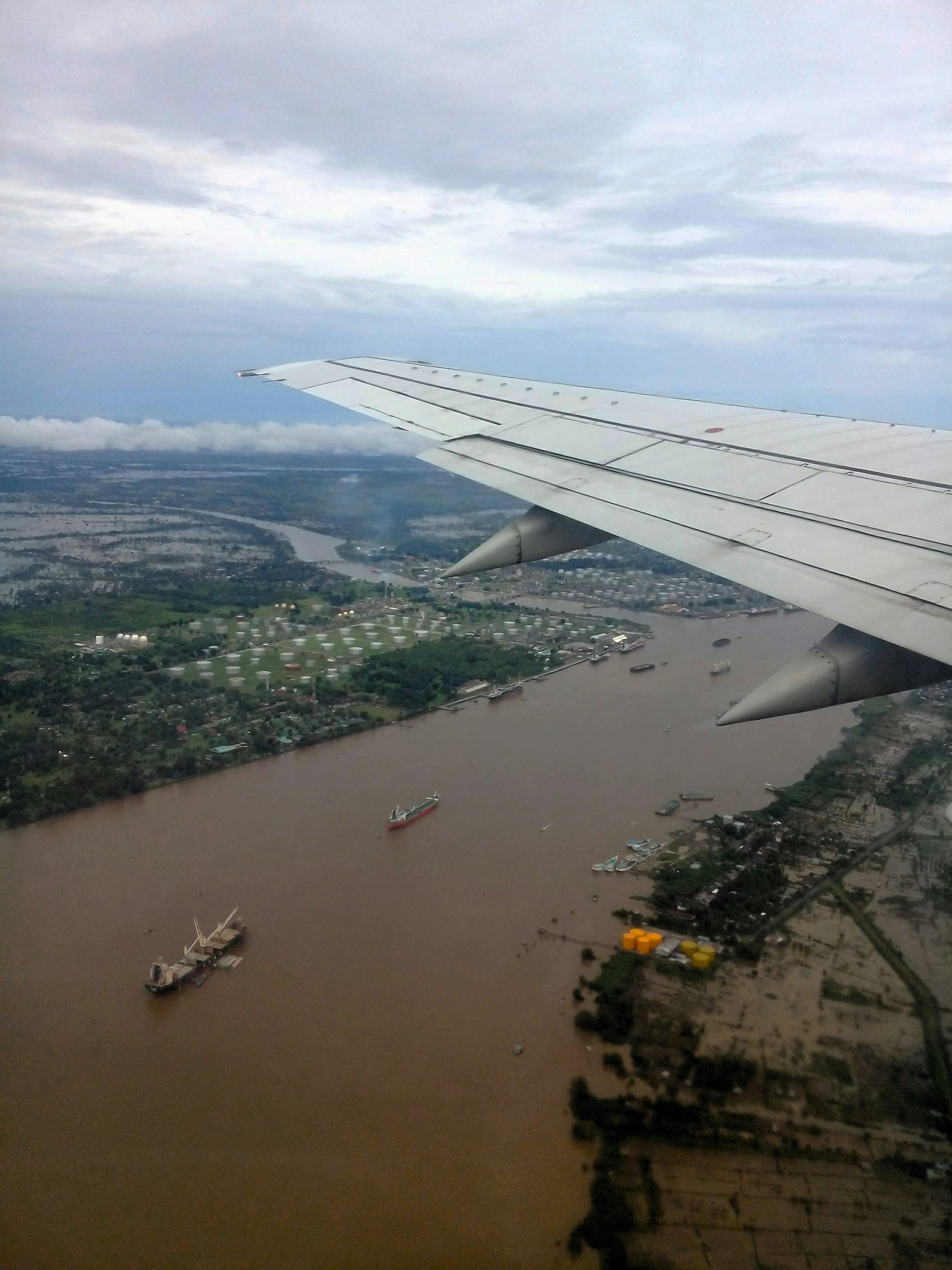 Airplane wing stretches across the frame. A muddy river winds through the riverbank landscape beneath an overcast sky.