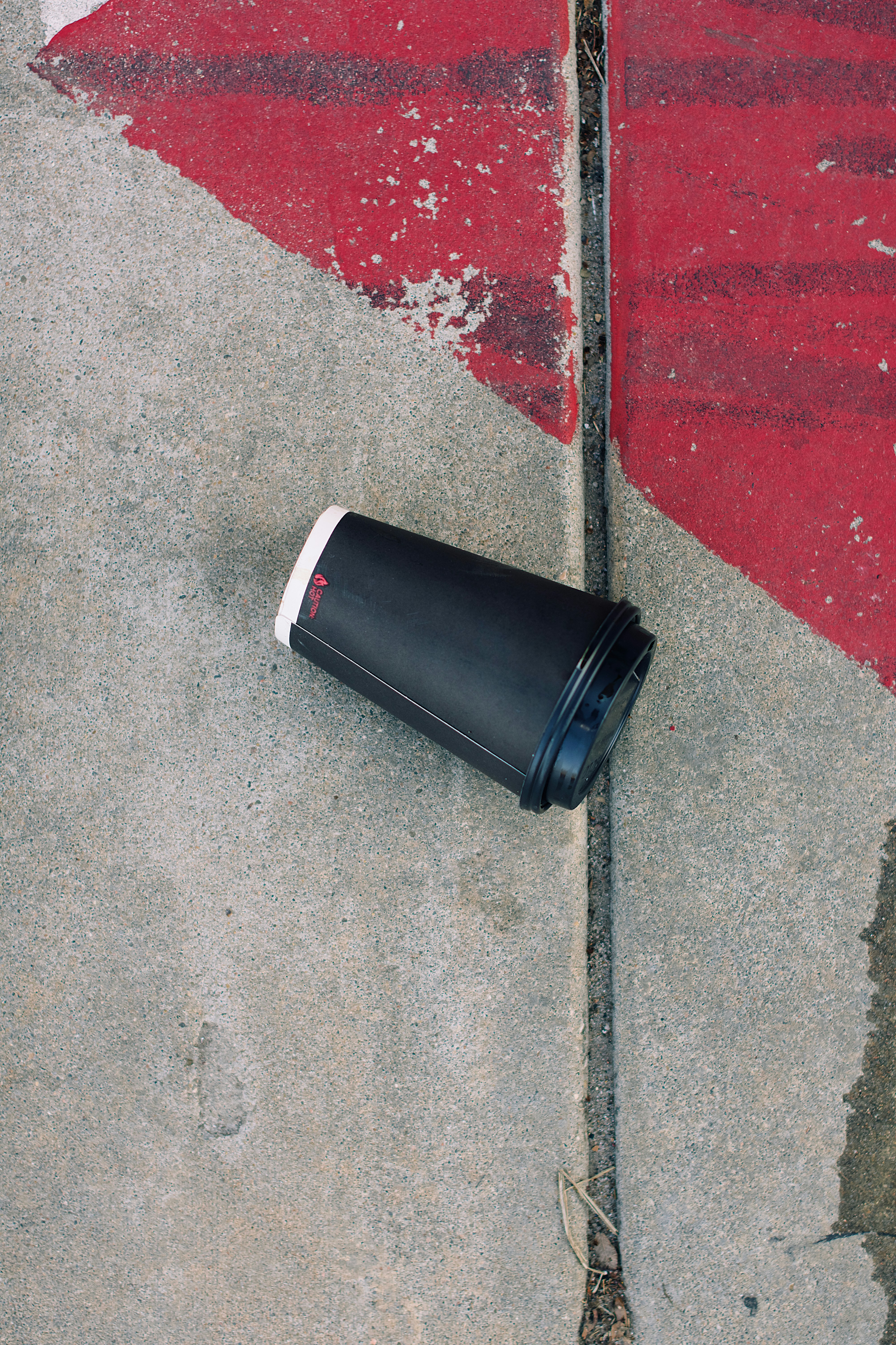 A discarded black coffee cup lies on a concrete surface, juxtaposed with vibrant red markings. The scene highlights urban neglect and the passage of time.