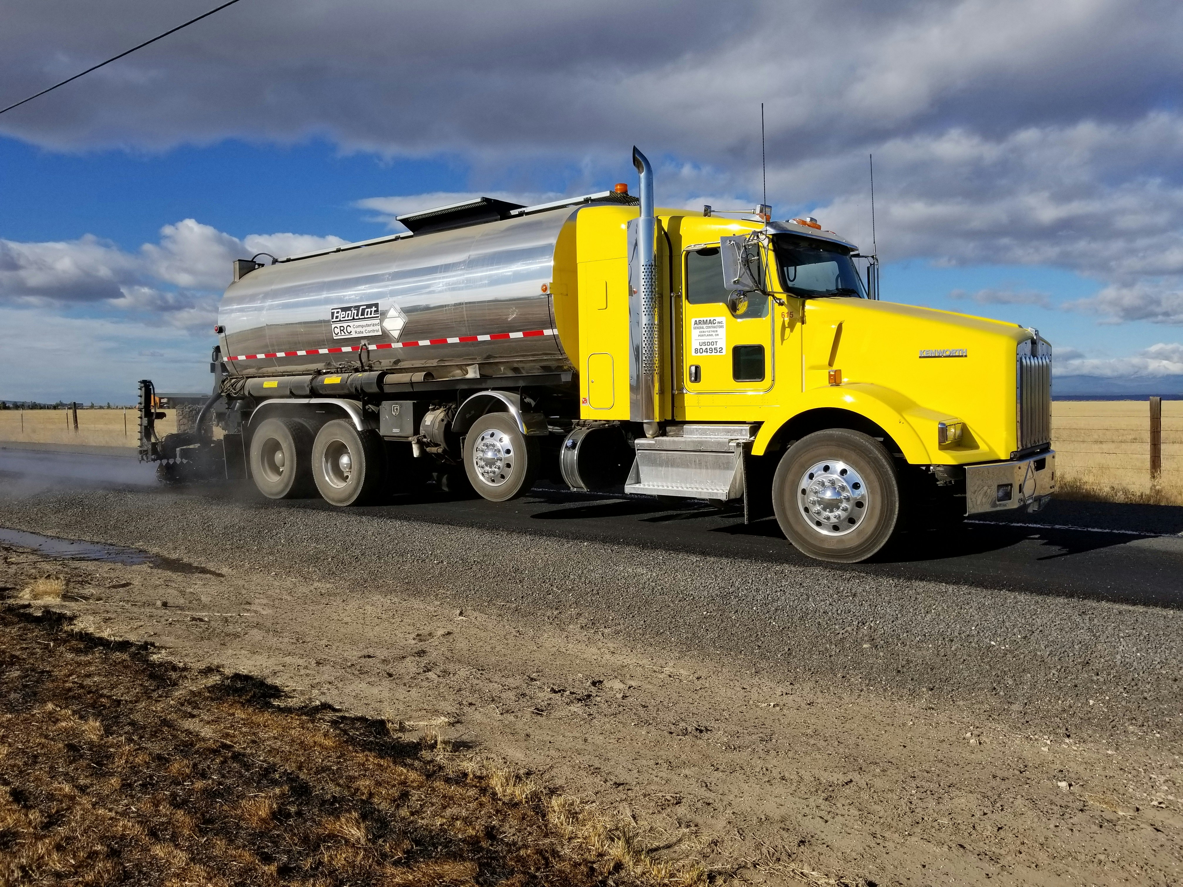 Yellow tanker truck applying emulsion oil on a rural road under a partly cloudy sky.