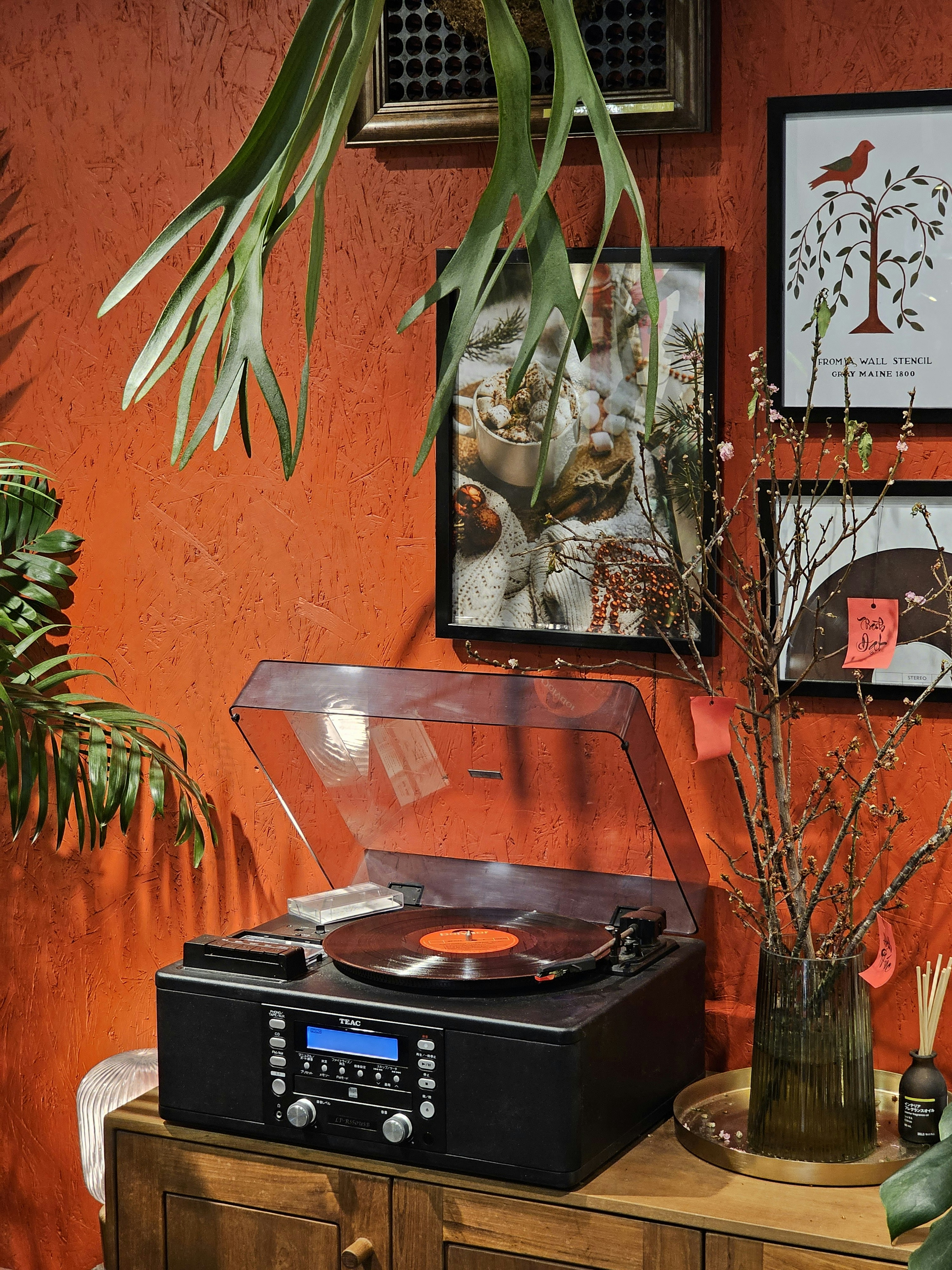 A vinyl turntable sits on a wooden cabinet against a textured orange wall, flanked by leafy plants and framed artwork.