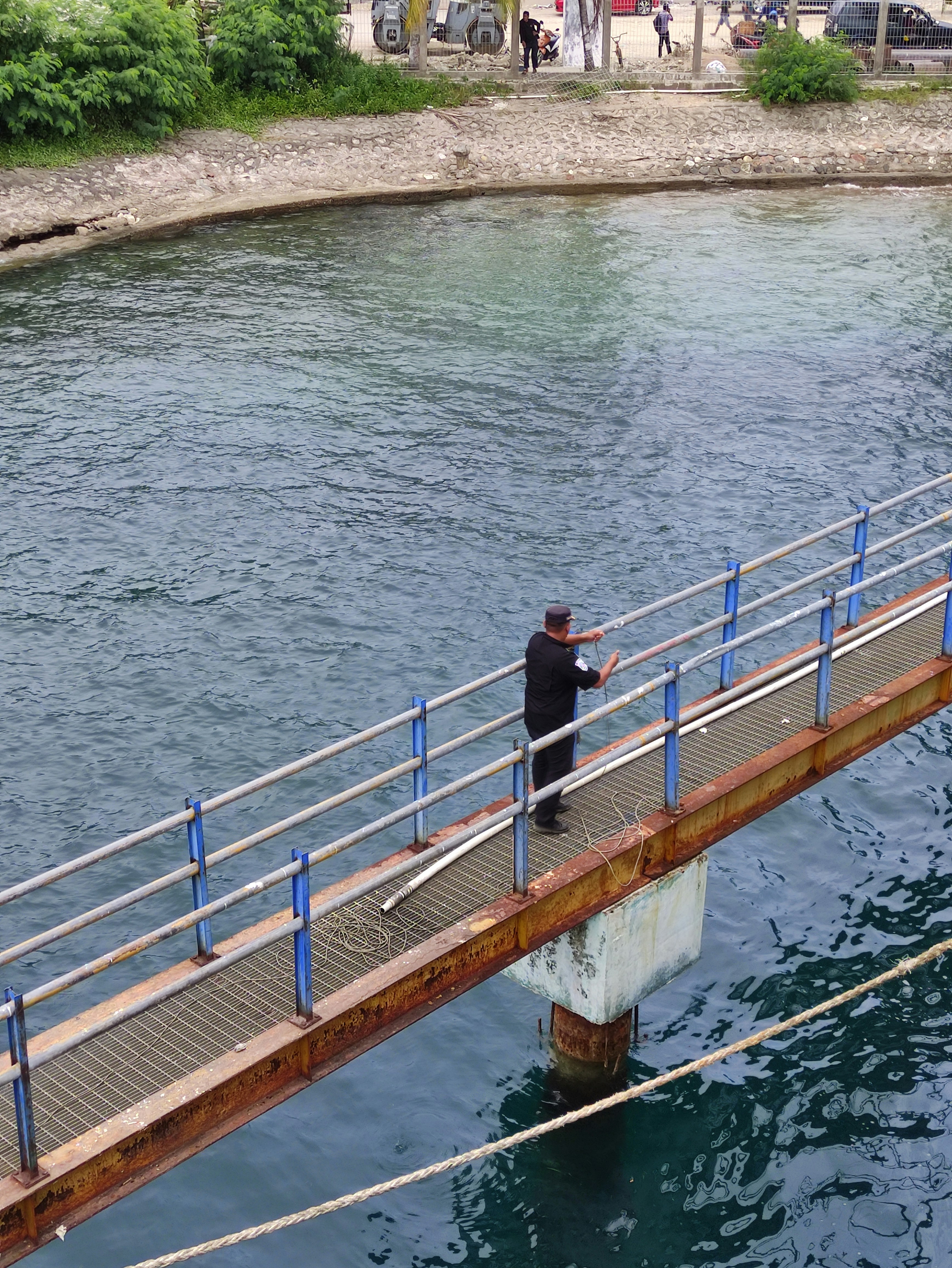 A solitary man stands on a rust-streaked pier as it juts over calm blue-green water, with a stone embankment and trees in the background.