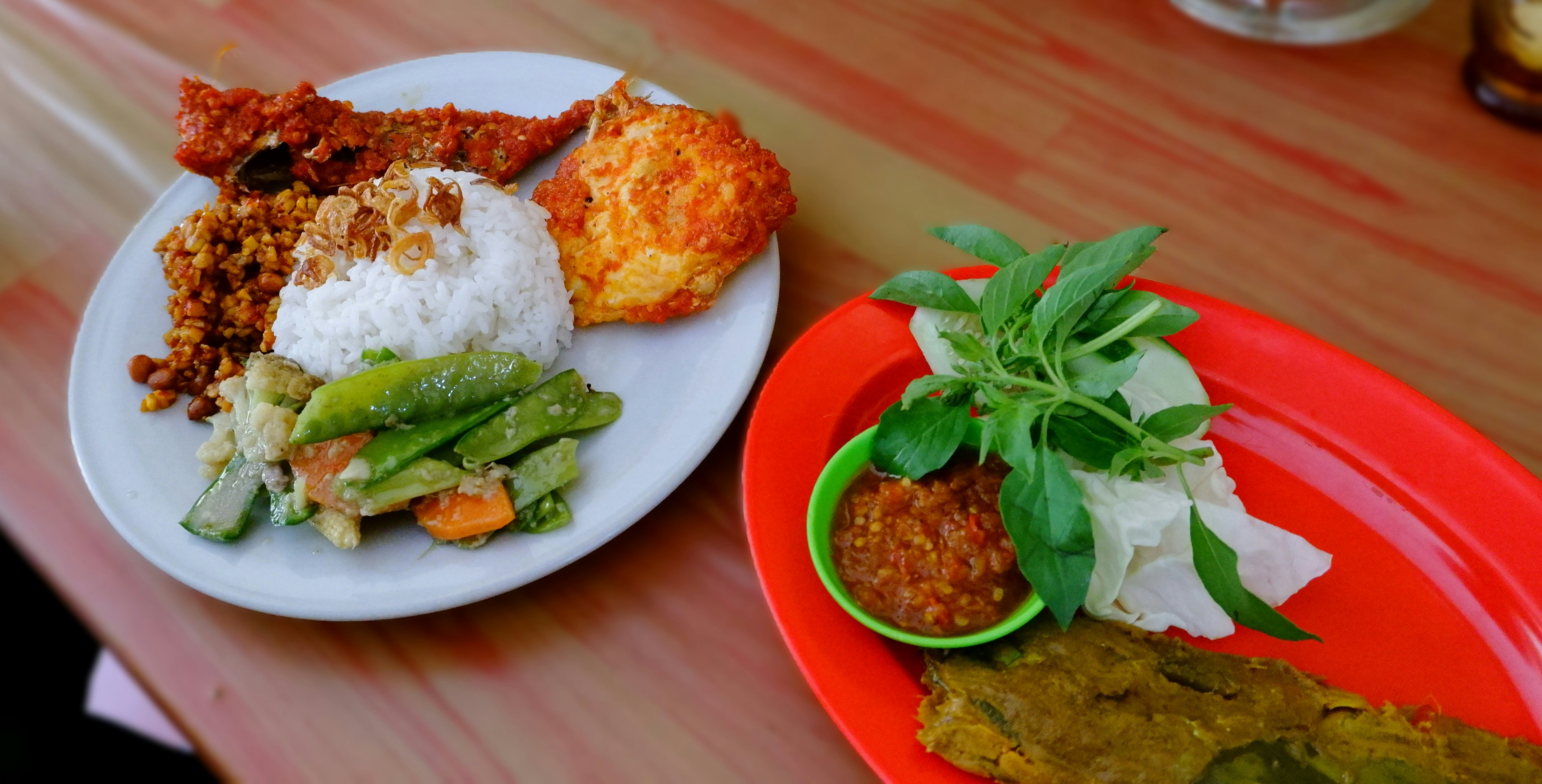 A vibrant plate featuring rice, assorted vegetables, and spicy condiments, showcasing a variety of textures and colors. The meal is served alongside a green herb garnish.