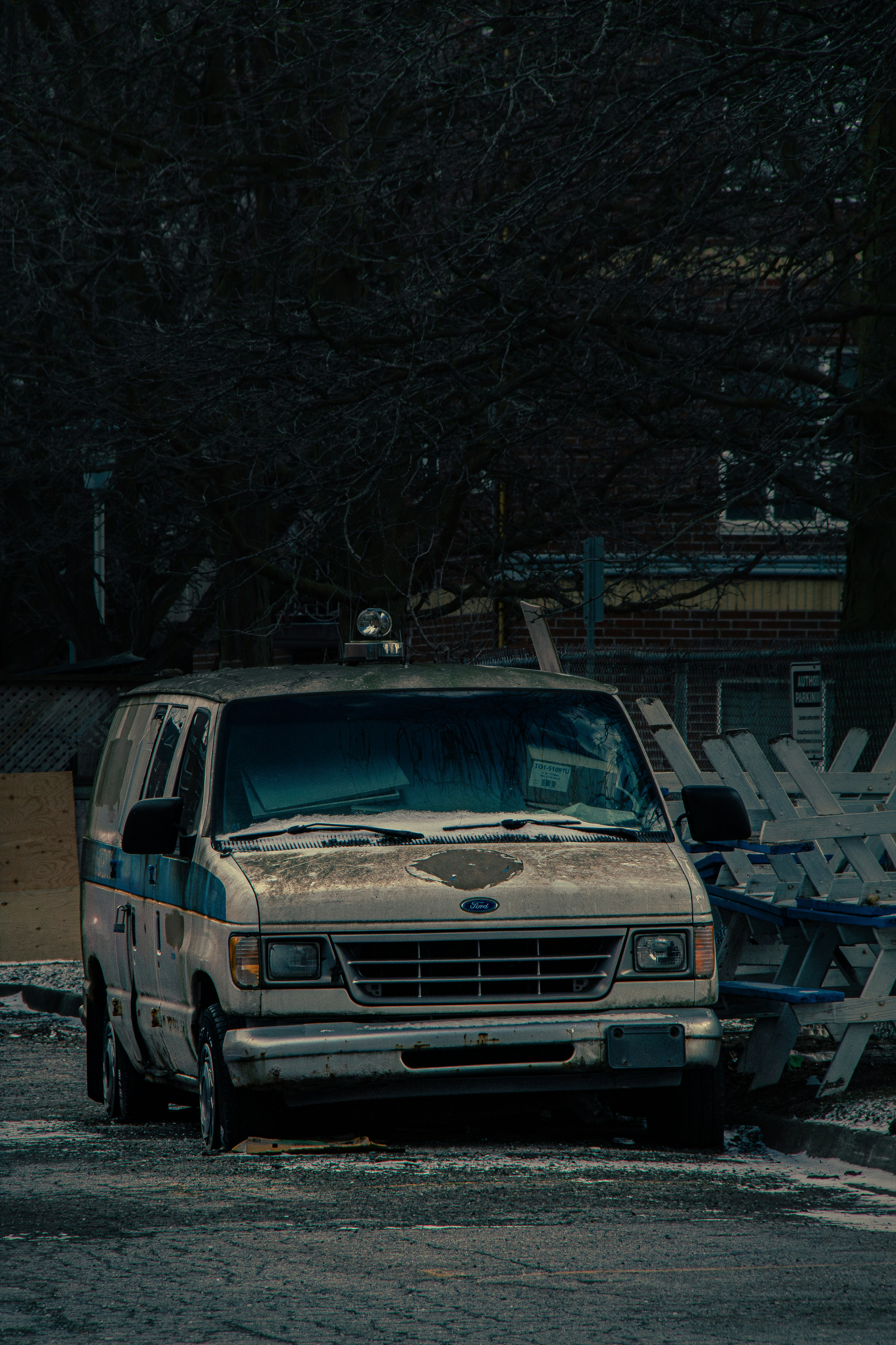 Weathered van under bare trees in a dim, debris-strewn lot