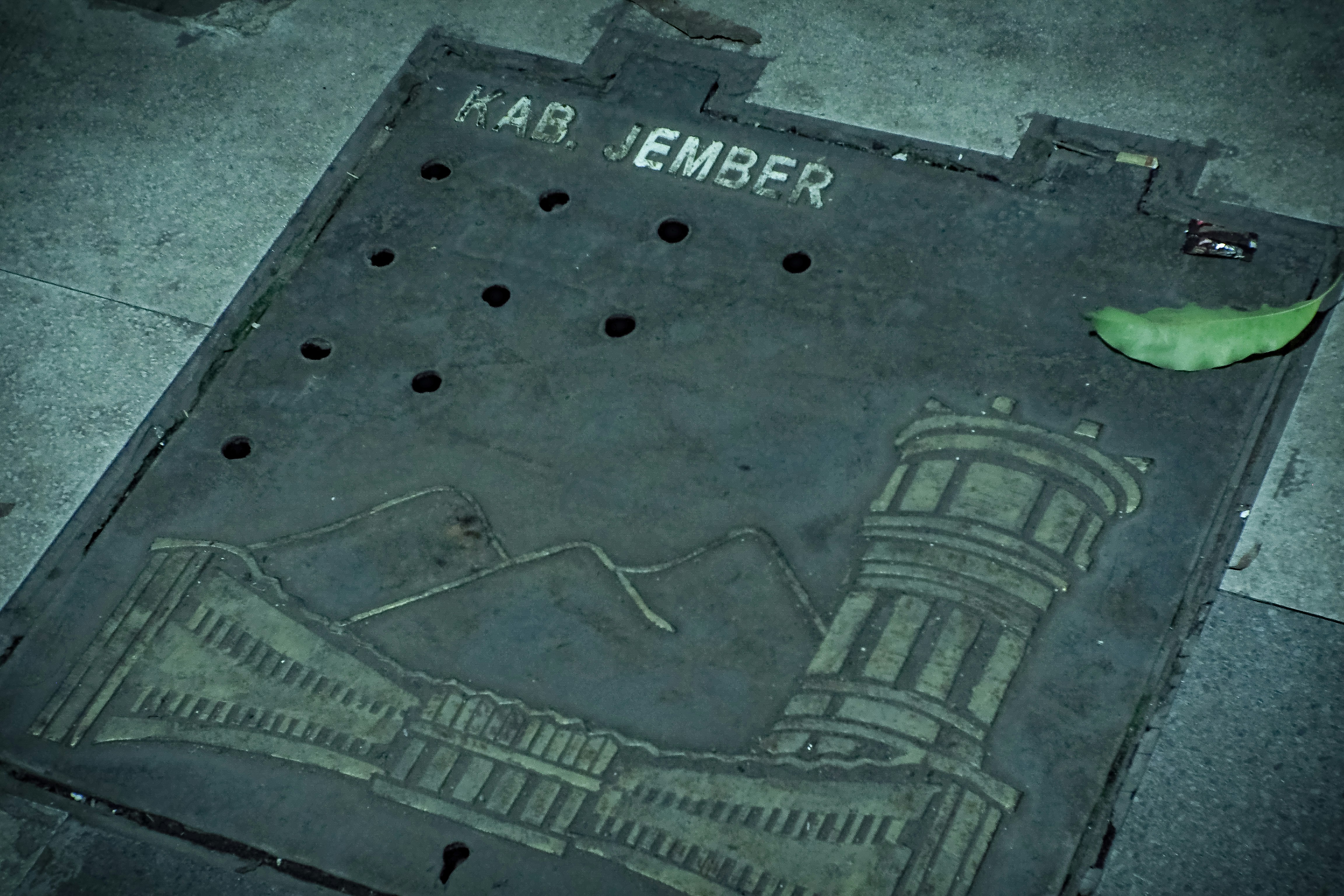 Decorative manhole cover featuring the name 'Kab Jember' and an intricate design of a tower and mountains. A single green leaf rests atop the cover.